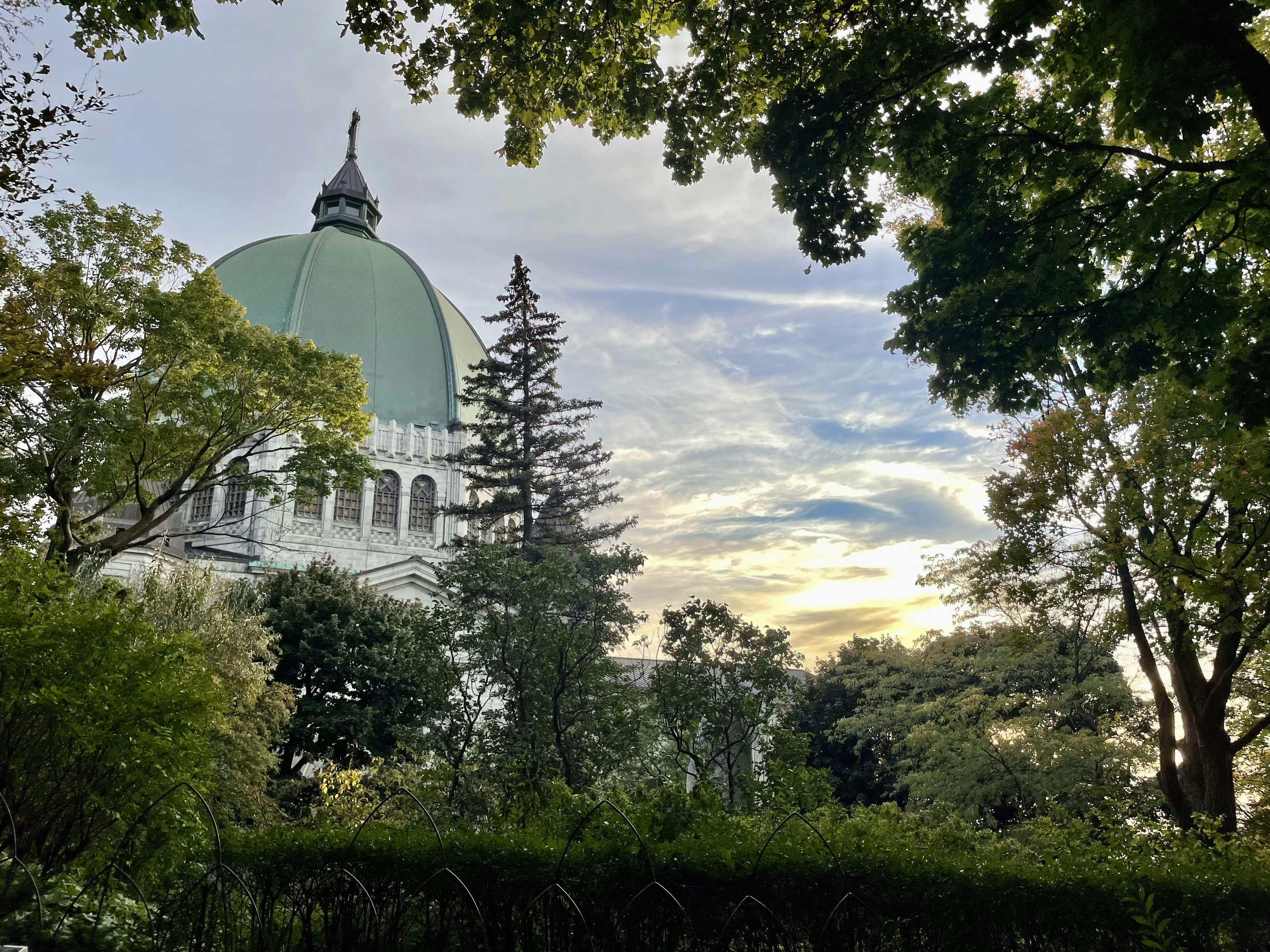 Green-domed building peeking through lush trees under a dynamic sky.