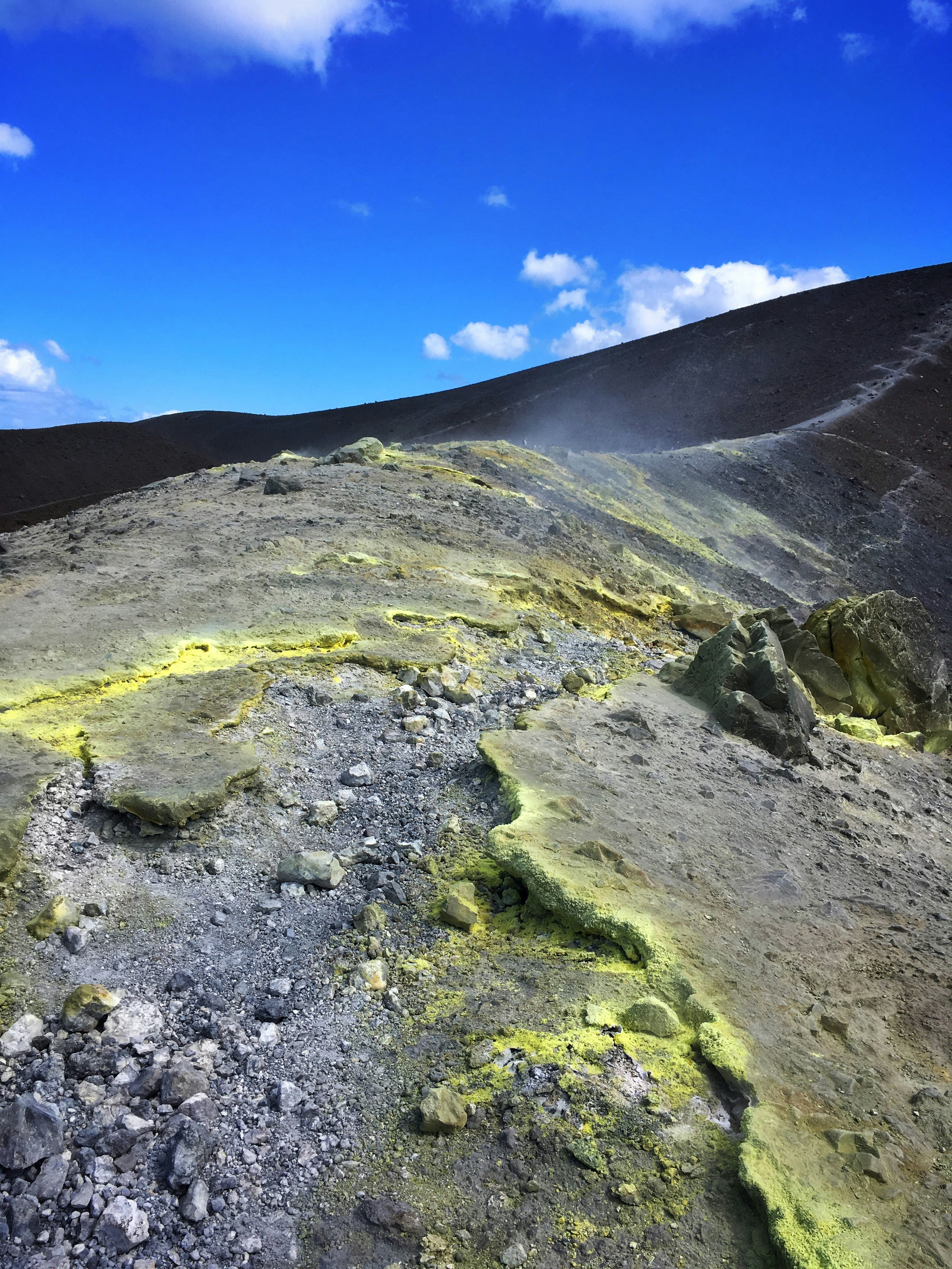 Vibrant yellow sulfur deposits contrast against a rugged volcanic terrain under a bright blue sky. The scene captures the raw beauty of geothermal activity.