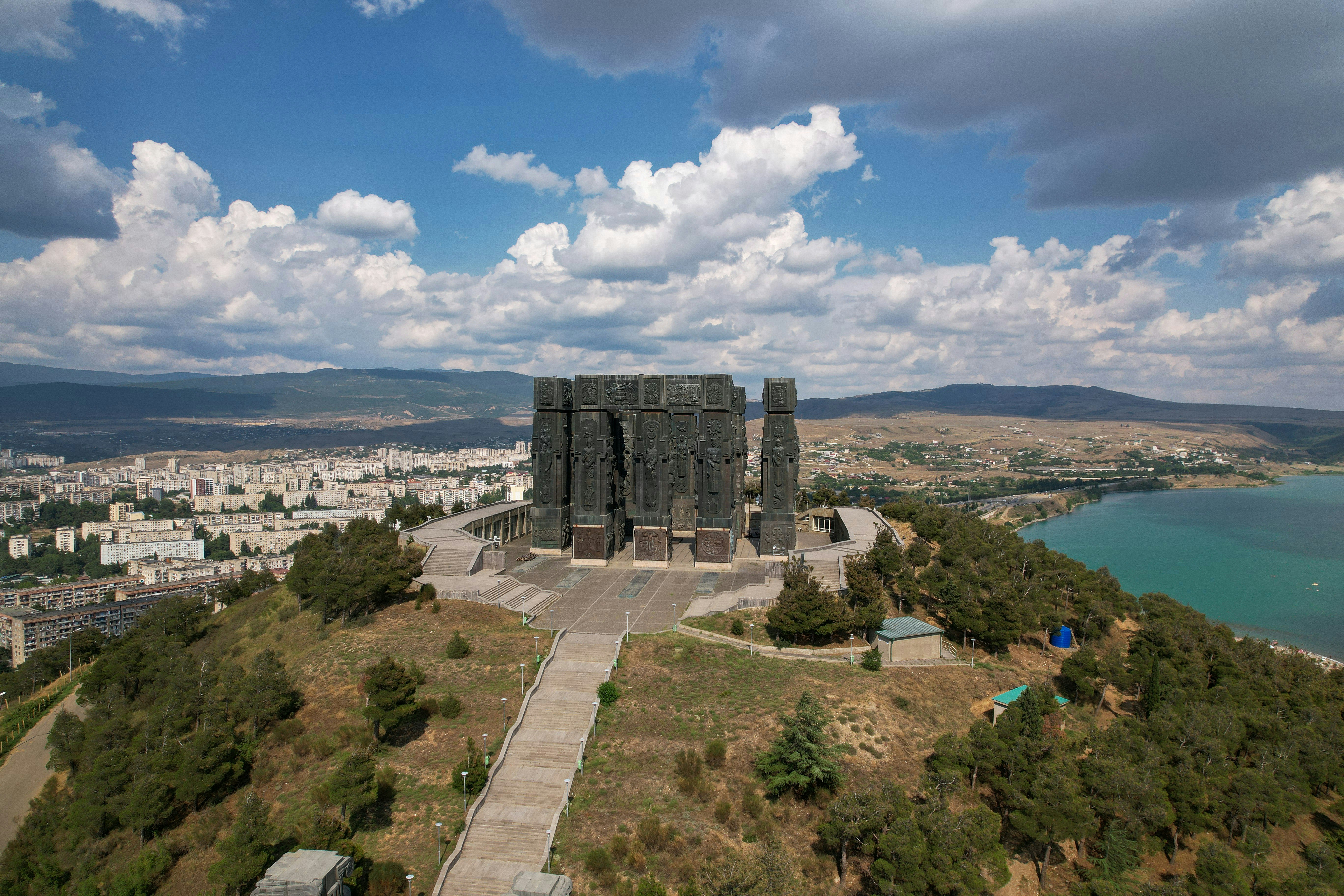 Aerial view of a monumental structure overlooking a scenic landscape, surrounded by greenery and water, showcasing the juxtaposition of nature and human achievement.