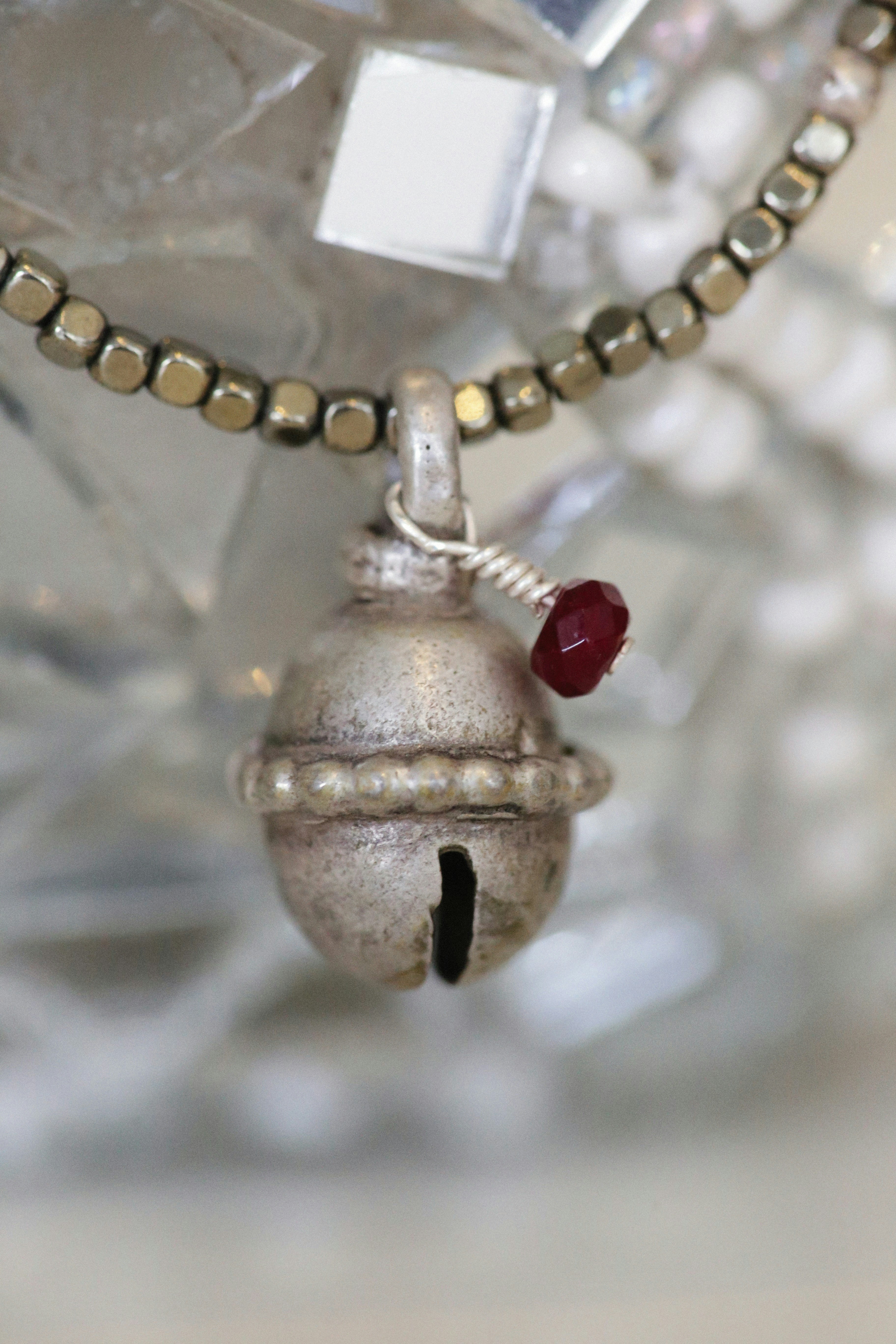 Intricately designed silver bell pendant adorned with a small ruby bead, suspended on a delicate chain against a reflective background.