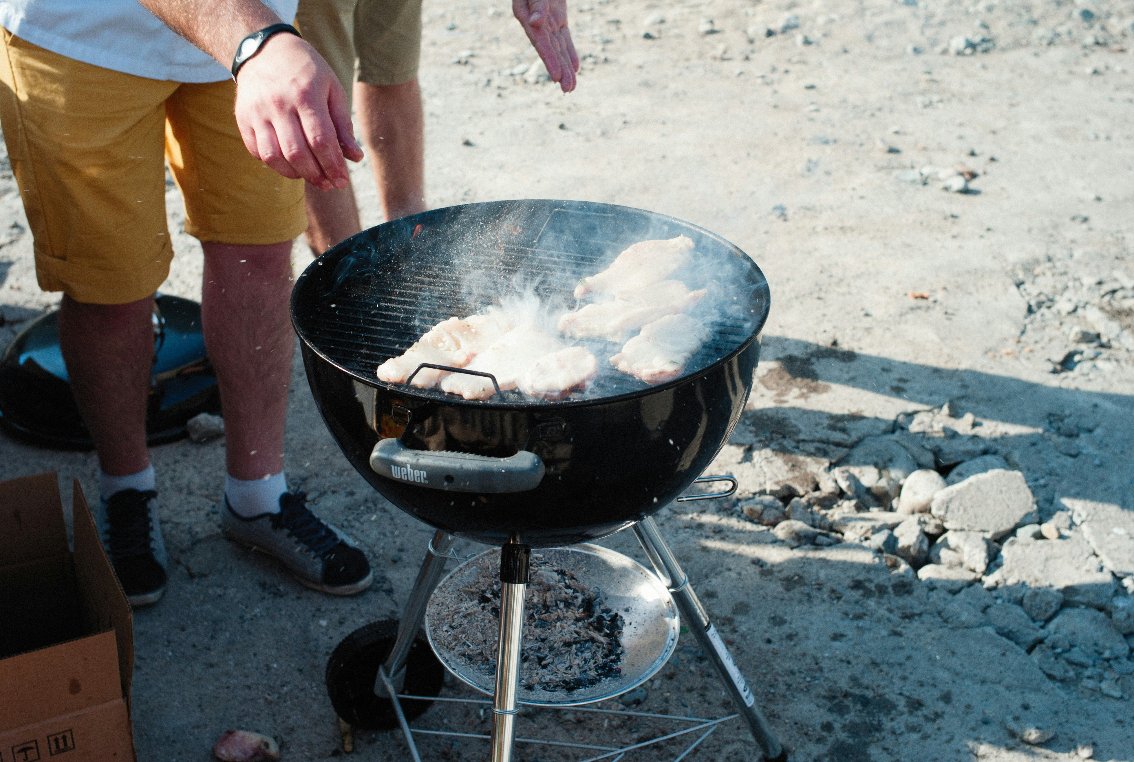 Hands grilling chicken on a charcoal barbecue at a casual outdoor gathering. Smoke rises as the food cooks over the open flame.