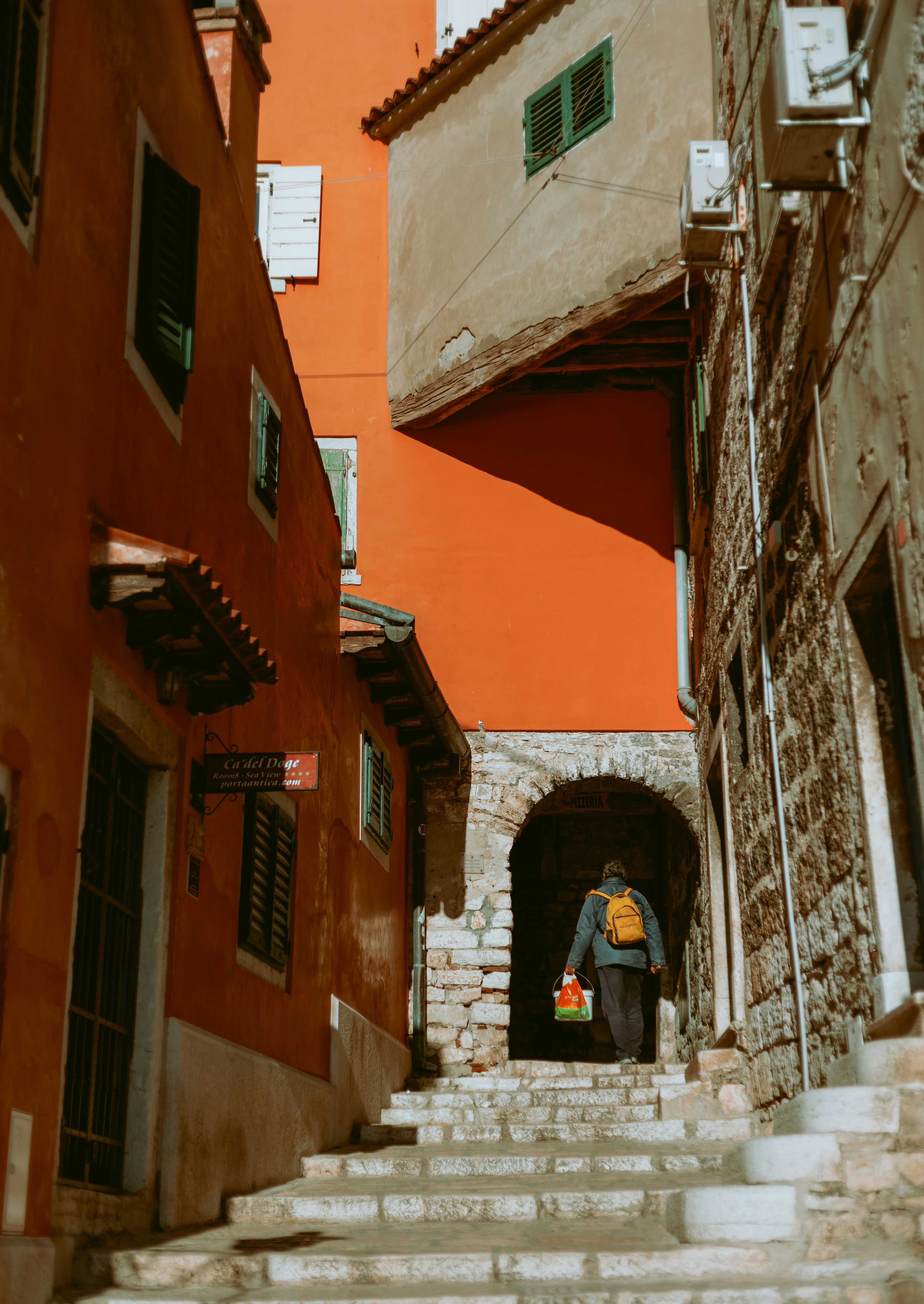 Person walking down a narrow stone staircase in a colorful alley, flanked by rustic buildings with vibrant orange and earthy tones.