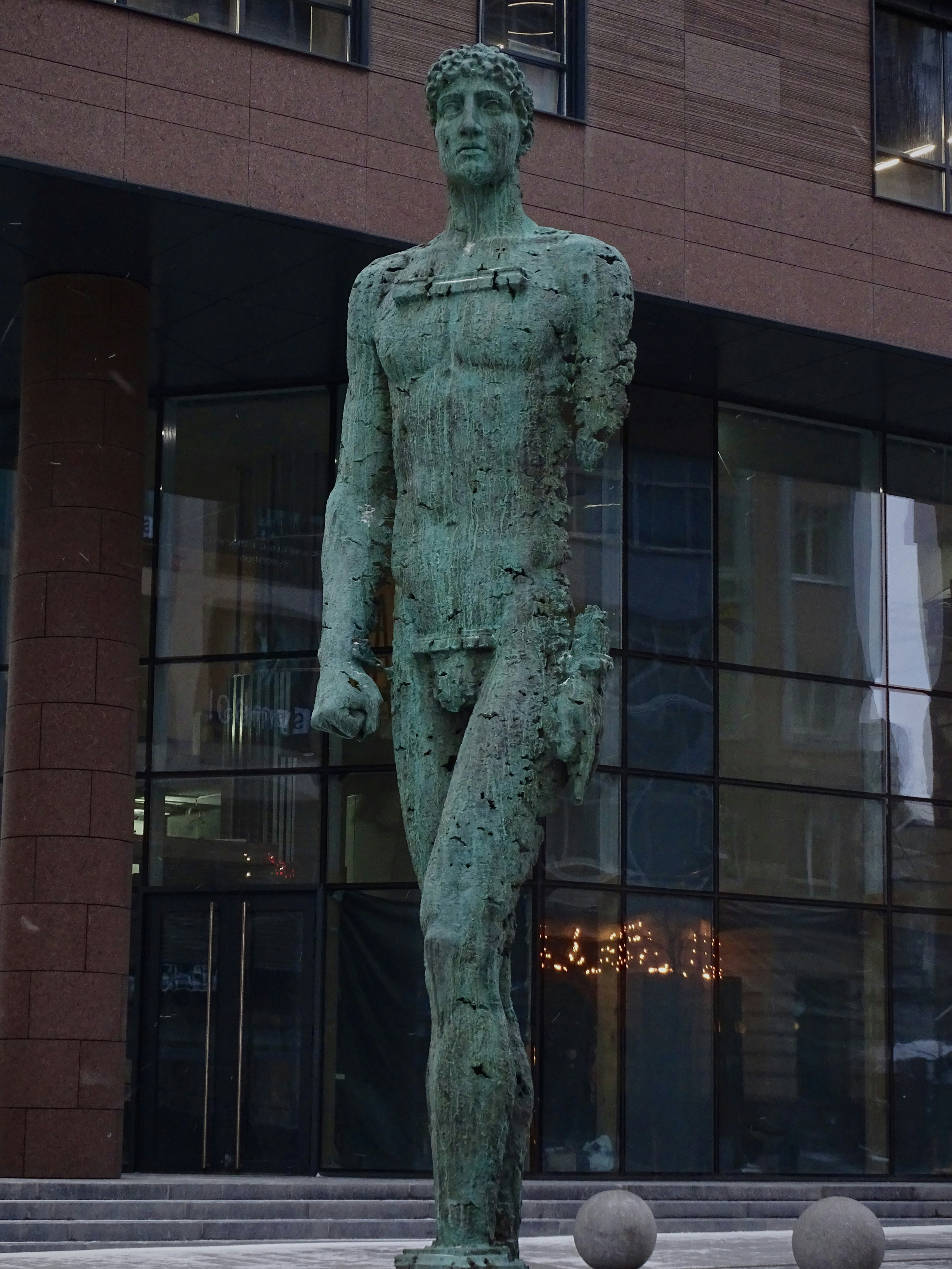 Bronze statue of a tall, slender figure standing in front of a modern building, showcasing intricate details and a weathered patina.