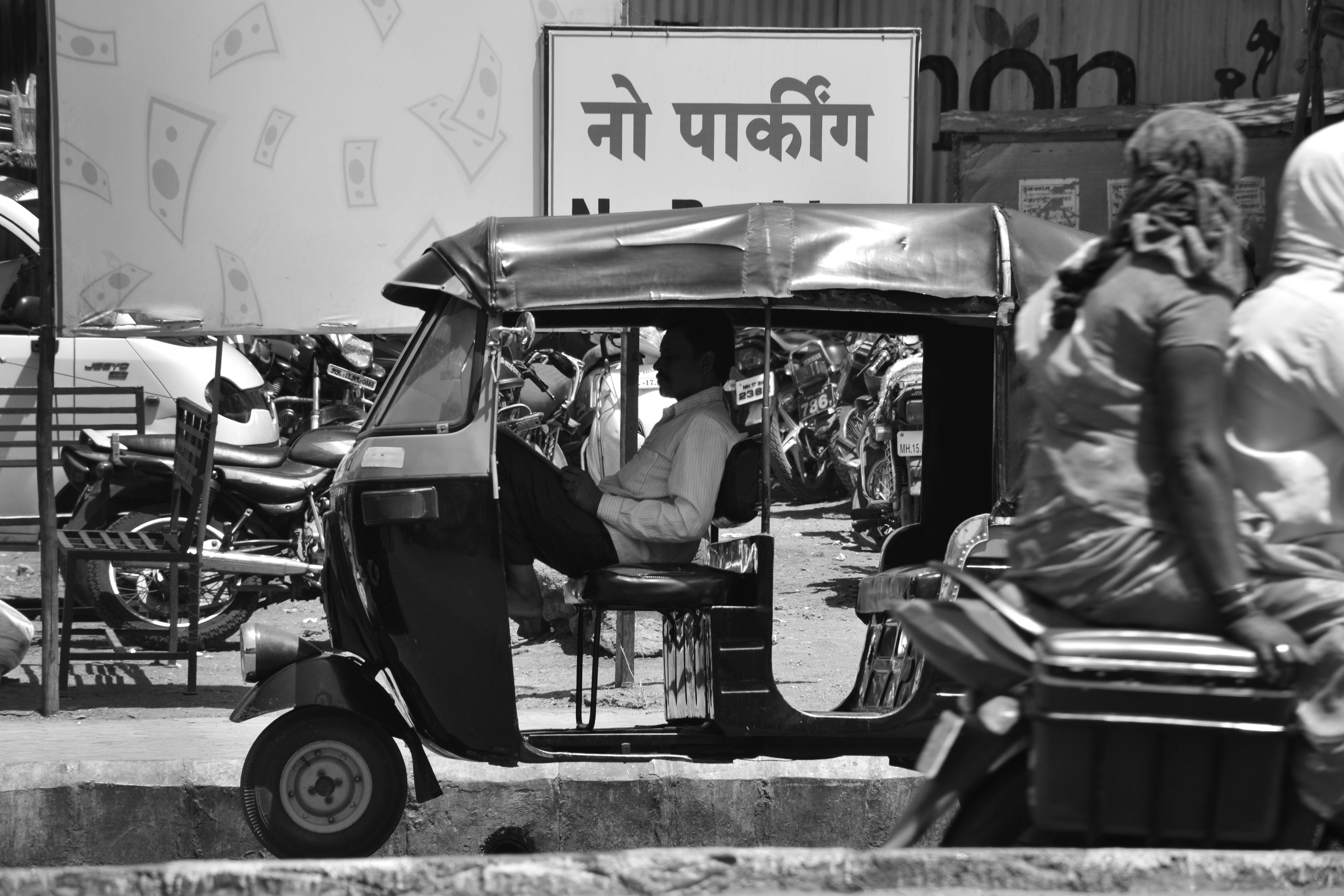 A rickshaw driver sits calmly in his vehicle amidst a bustling street scene, framed by parked motorcycles and a sign in the background. The monochrome tones emphasize the urban environment.