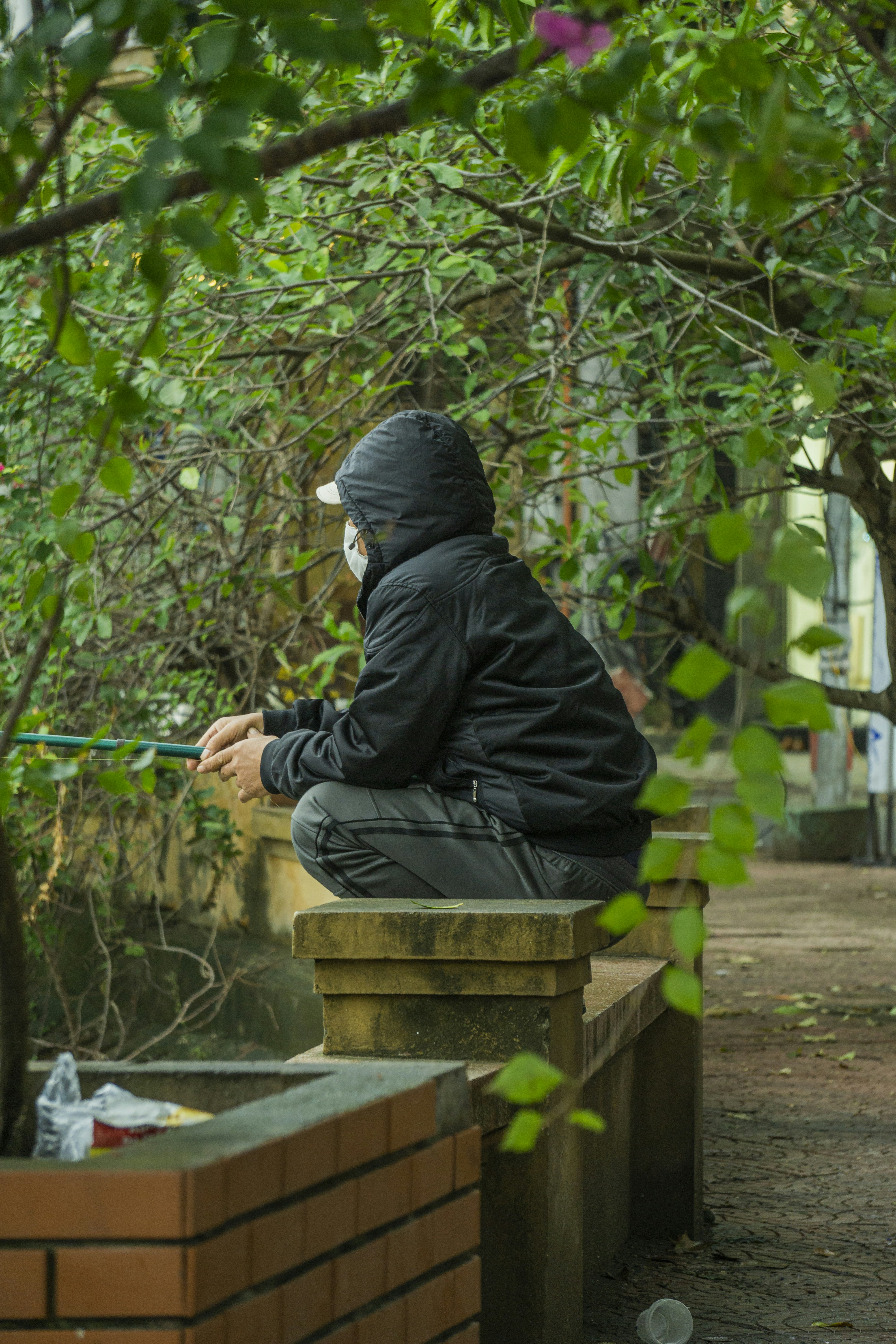 Individual in a black jacket sitting on a stone ledge, engaged in quiet contemplation near a lush, green setting.