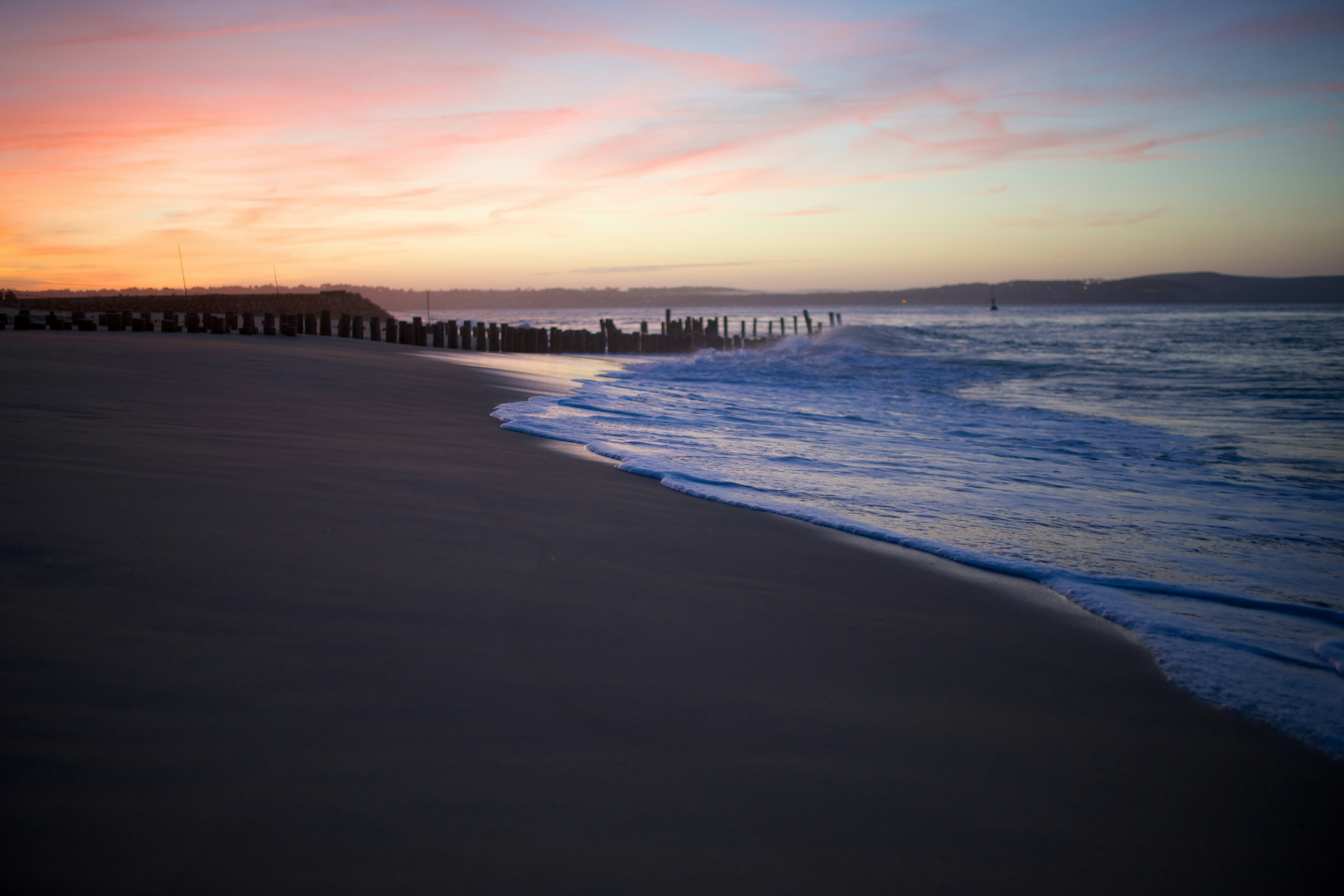Sunset beach with weathered pylons stretching into the calm sea. The sky glows pink and orange as waves lap the shore.