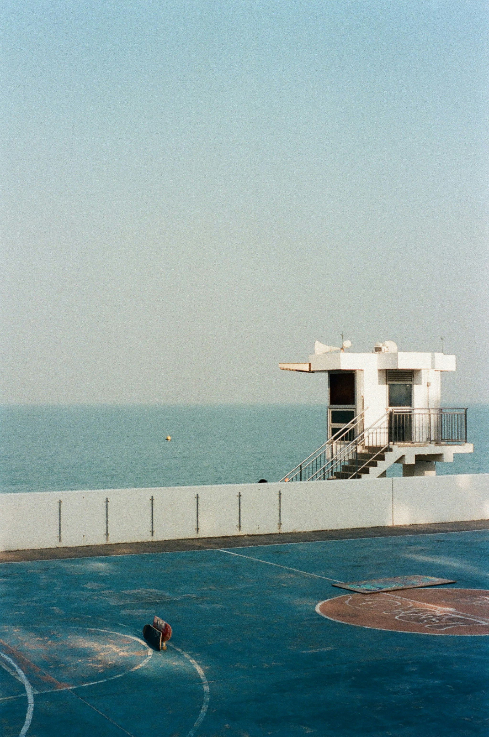 Lifeguard tower overlooking calm sea with a basketball court in the foreground. The scene captures a serene coastal atmosphere.