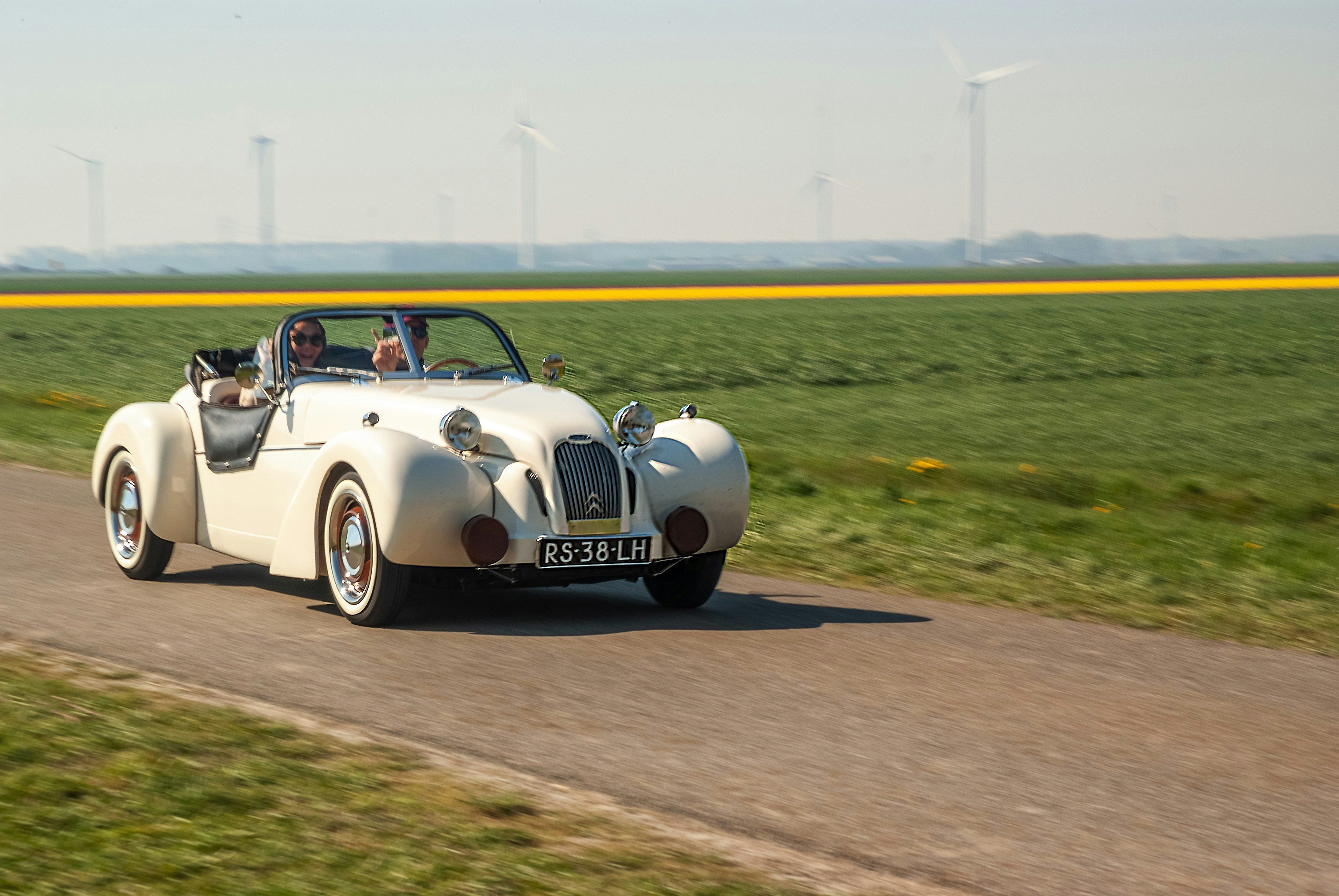Classic convertible car cruising along a rural road, surrounded by vibrant fields and wind turbines in the background.