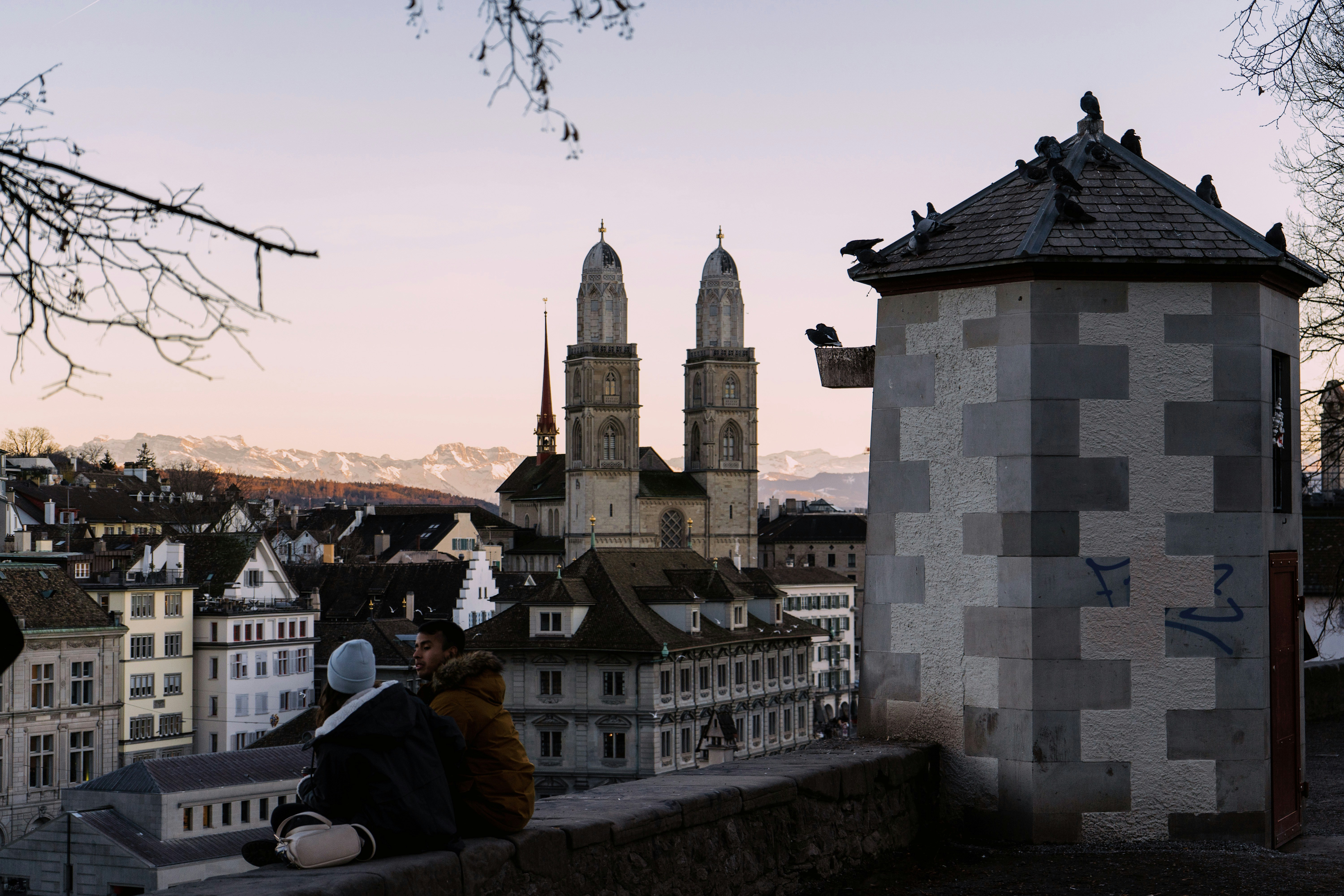 Couple seated on a stone wall, overlooking Zurich's skyline with historic architecture and distant mountains at twilight.