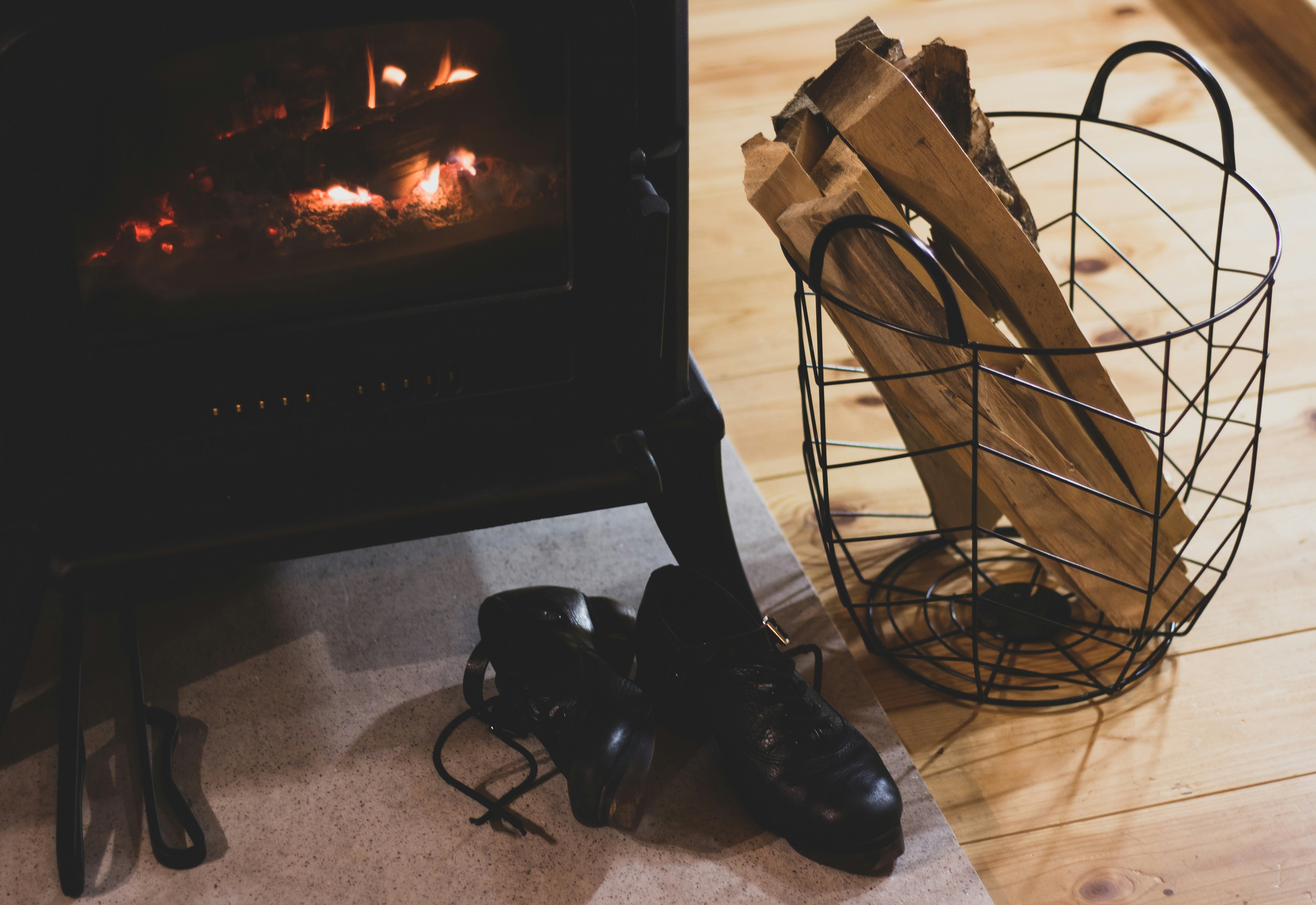 Cozy scene featuring a wood-burning stove with glowing embers, alongside a wire basket filled with firewood and a pair of black shoes resting on a rug.