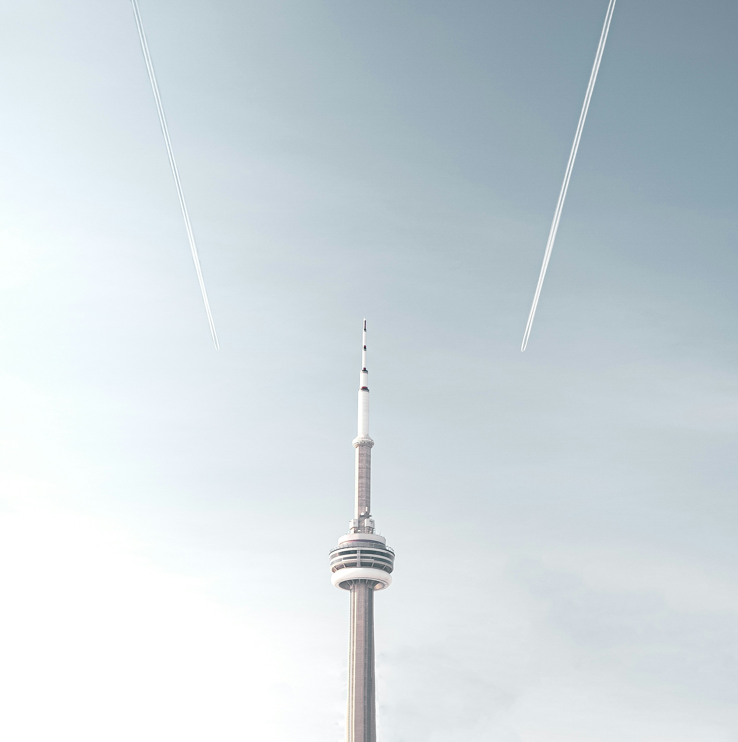 The CN Tower rises prominently against a pale blue sky, accented by two contrails that stretch across the horizon.