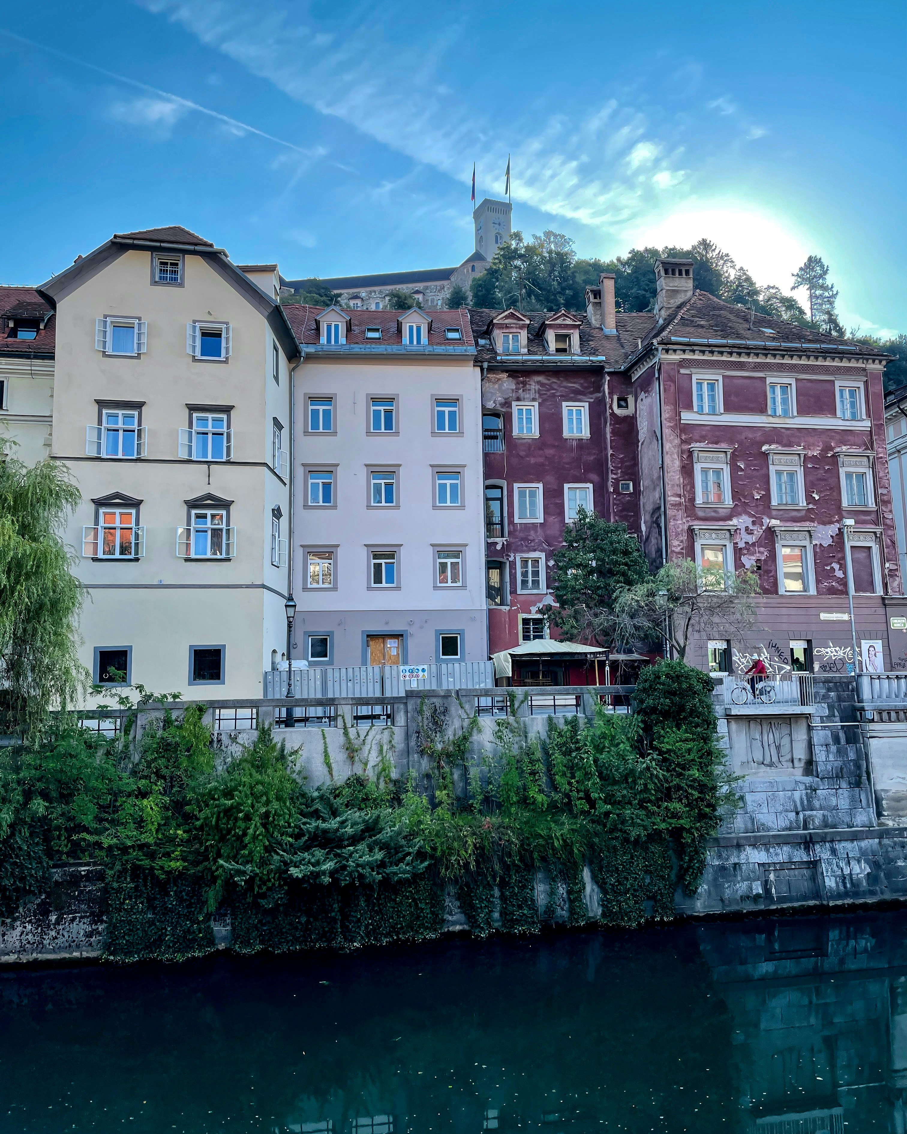 Colorful buildings line the riverbank, with a historic castle perched on a hill in the background. Lush greenery adds to the serene atmosphere.