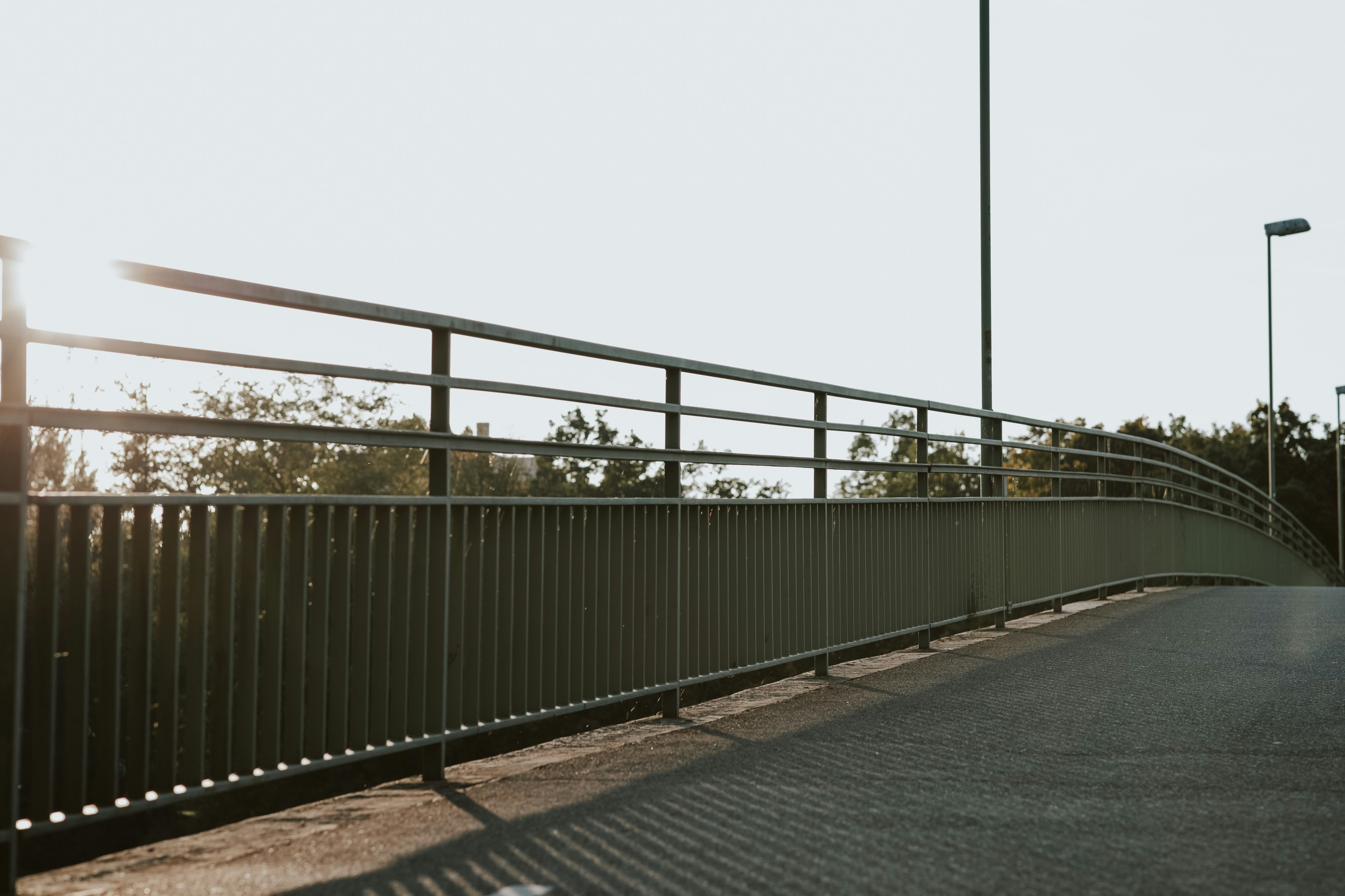 A gently sloping pedestrian bridge with a curved railing, bathed in soft sunlight, leading towards a lush green backdrop.