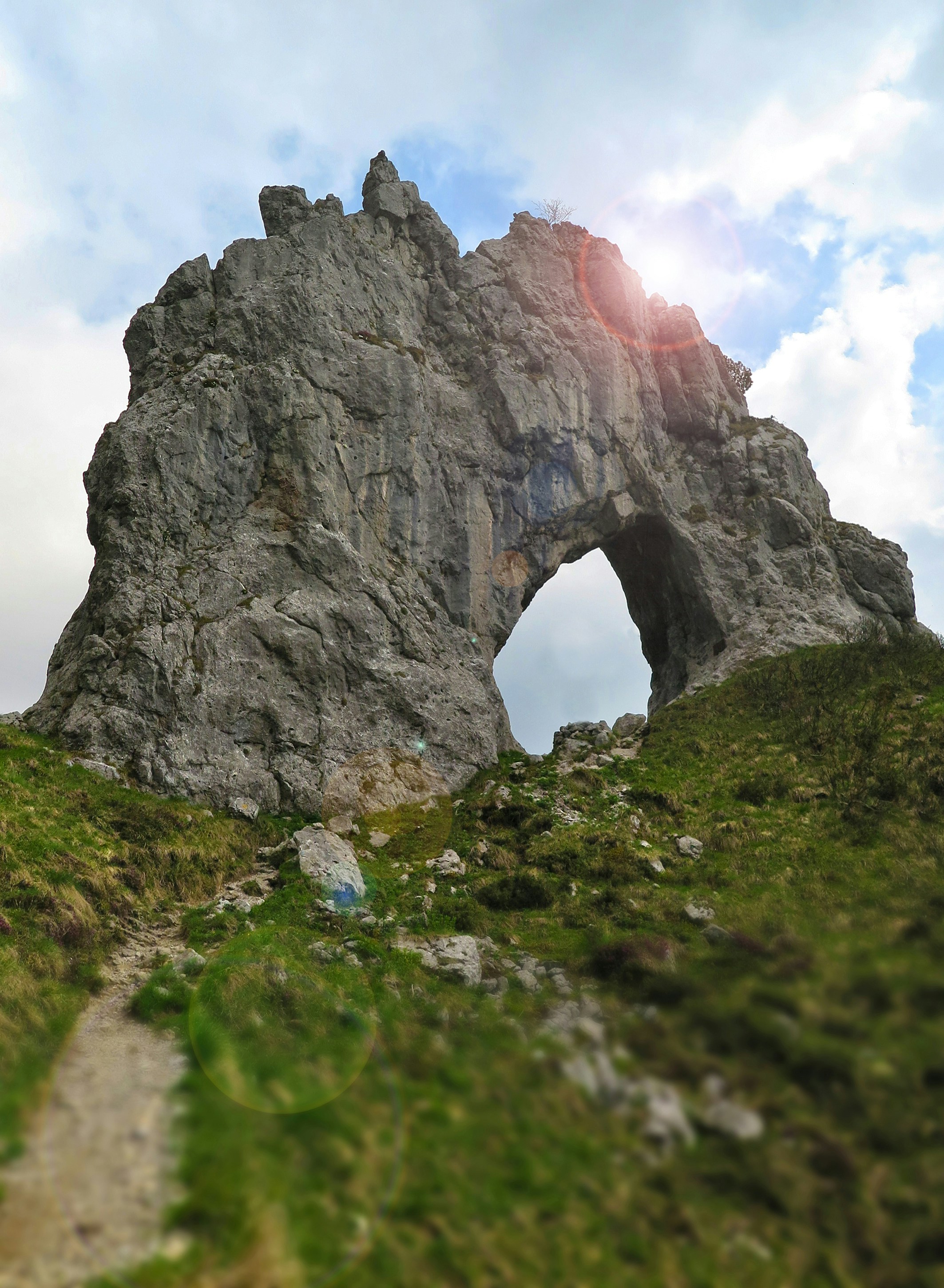 Rock arch rises from a grassy hillside in a landscape photograph, with a rocky path leading toward its opening and a sun flare at the top.