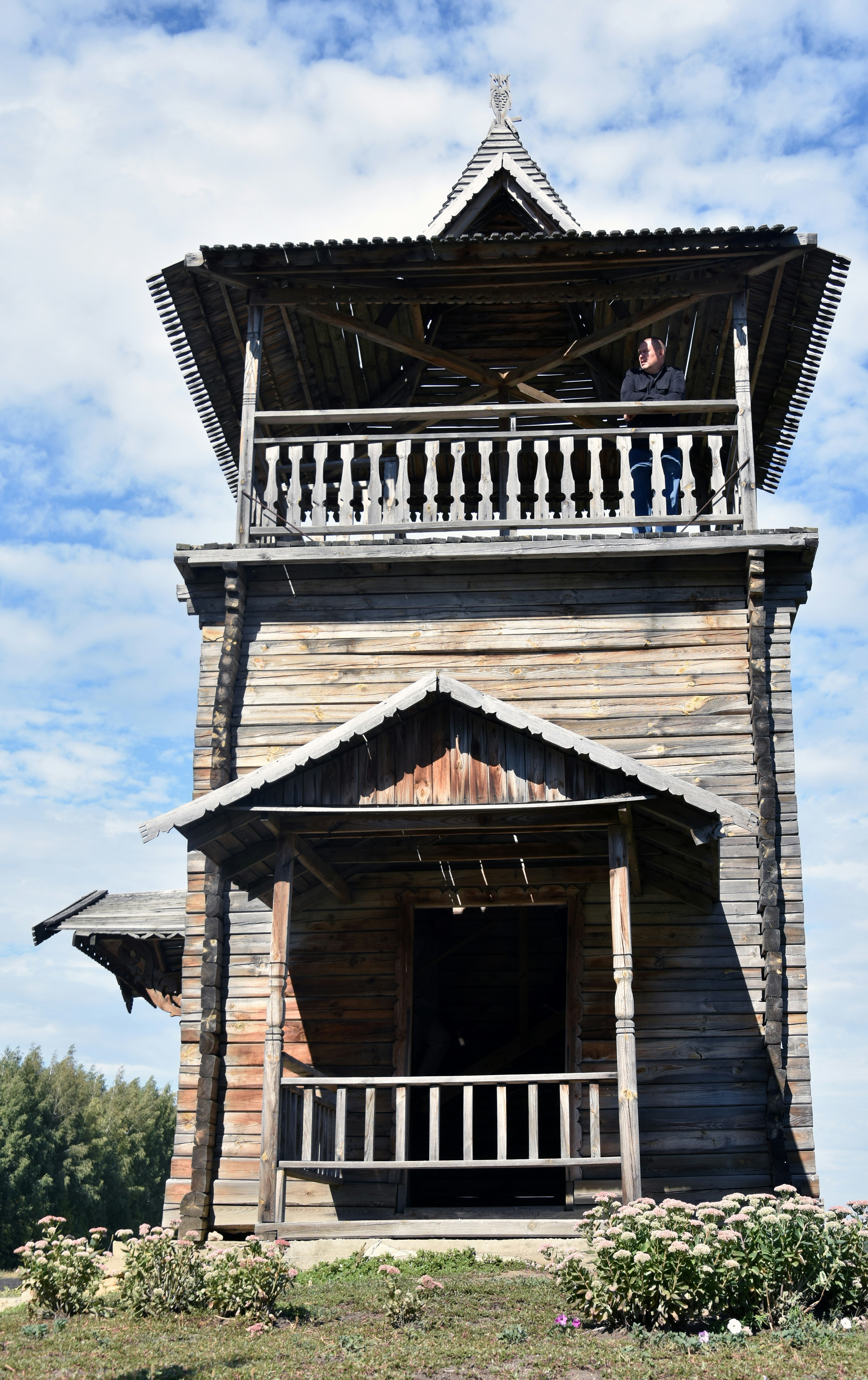 A rustic wooden tower stands tall against a cloudy sky, with a figure peering from its balcony, evoking a sense of history and solitude.