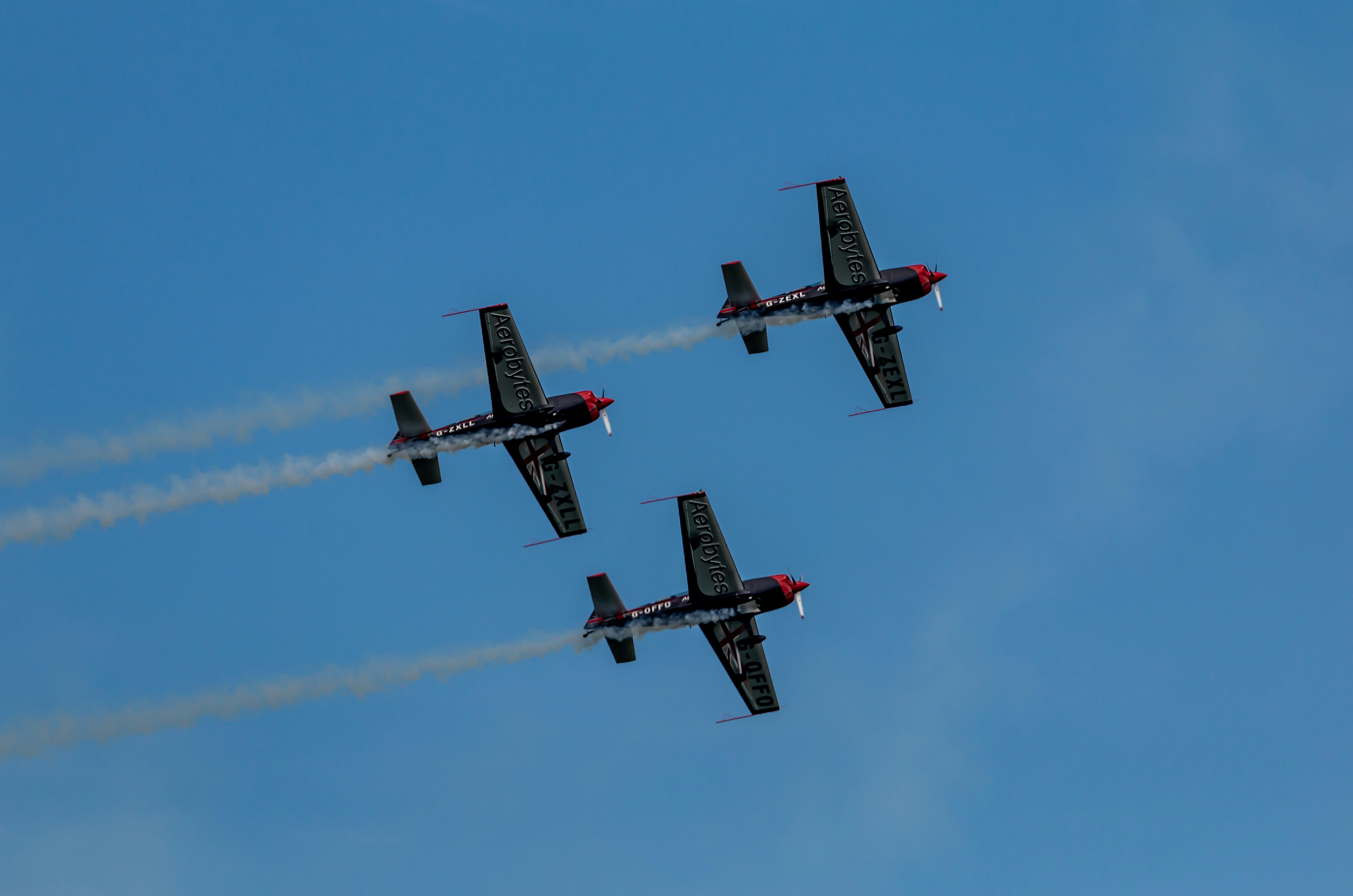 Three aircraft performing a synchronized aerial formation against a bright blue sky, leaving trails of smoke behind.