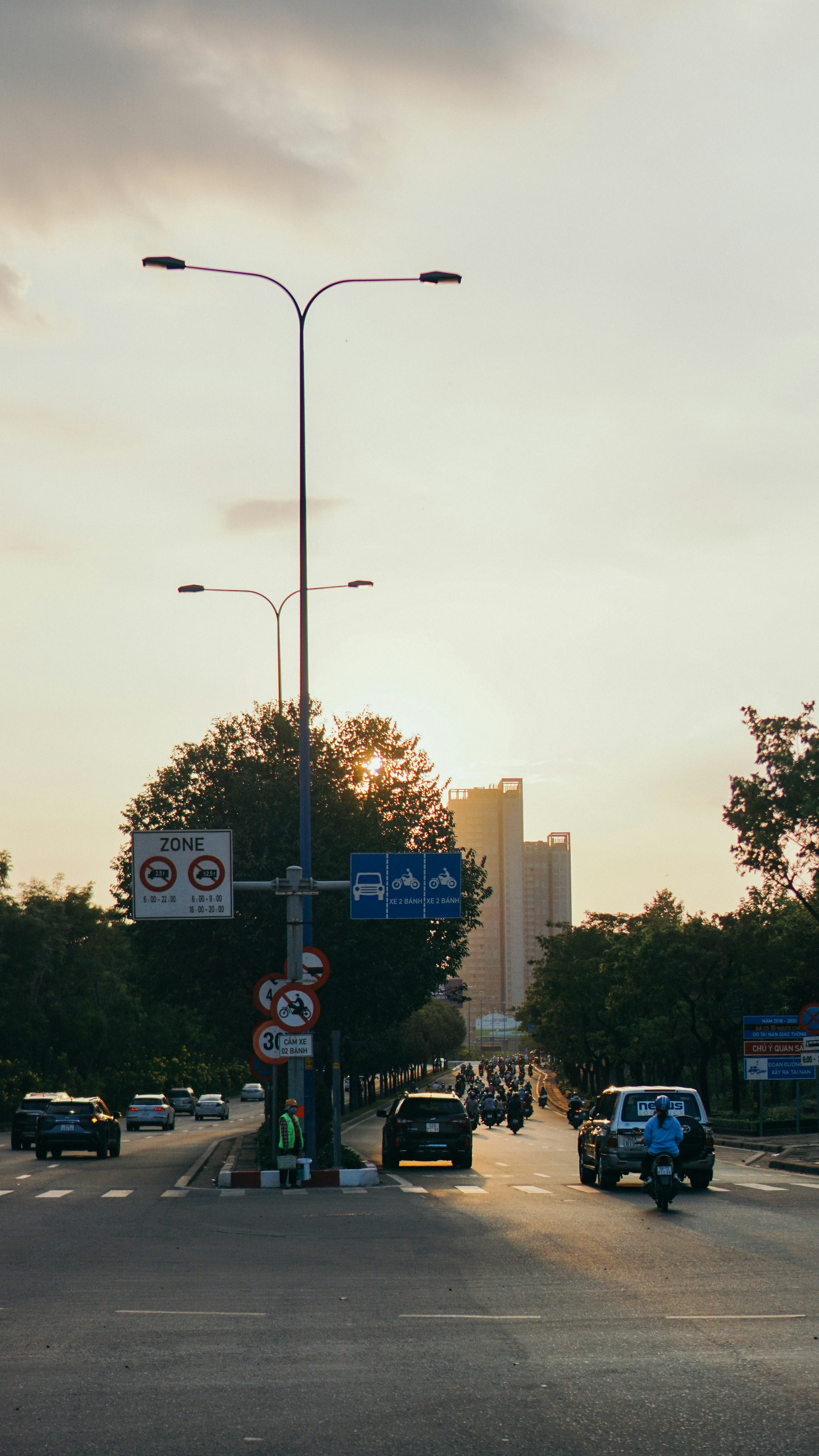 Traffic flows along a city street as the sun sets behind tall buildings, casting a warm glow. Road signs and streetlights frame the scene.