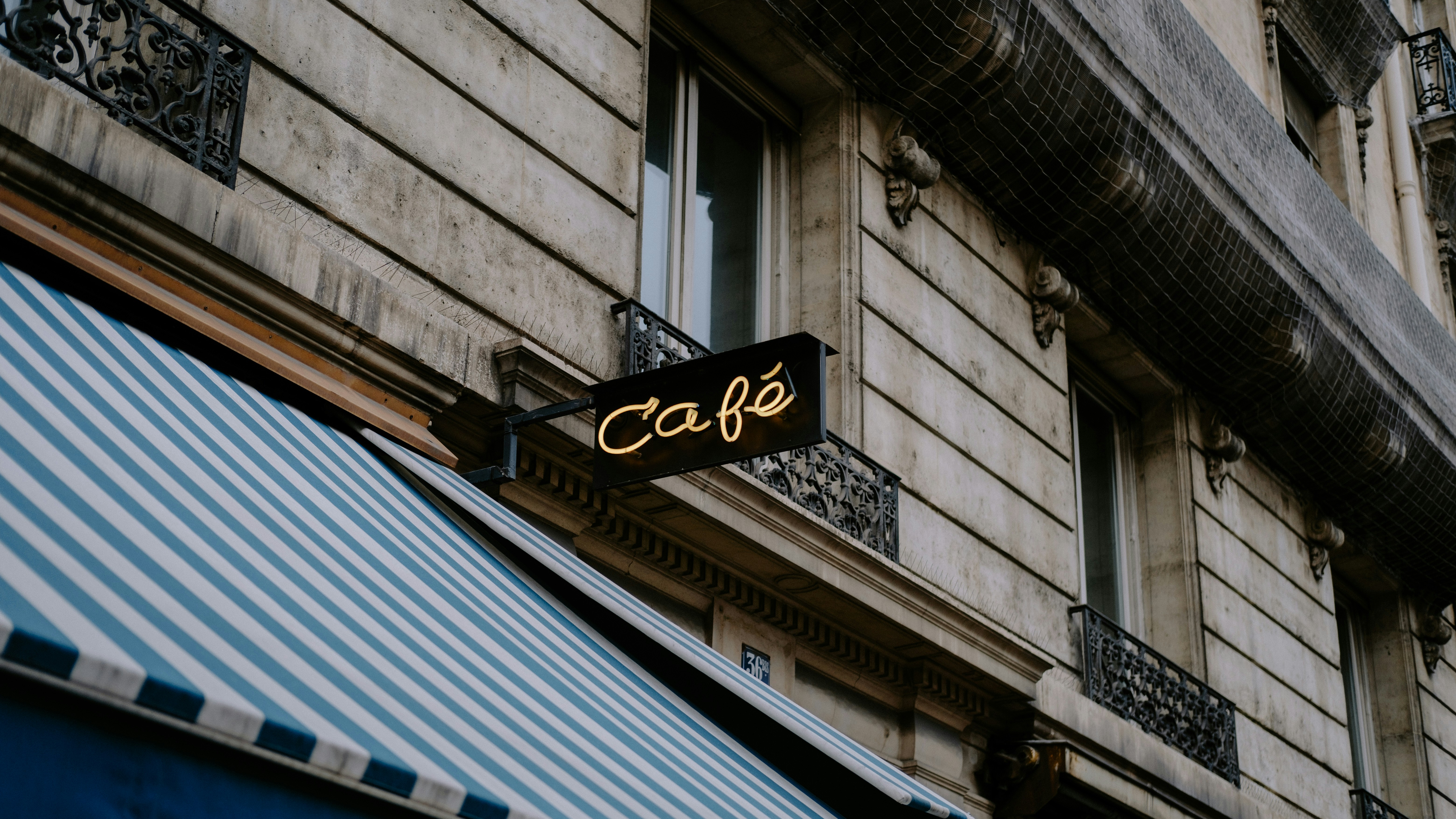 Illuminated café sign hanging above a striped awning on a classic building facade, showcasing Parisian architectural elegance.