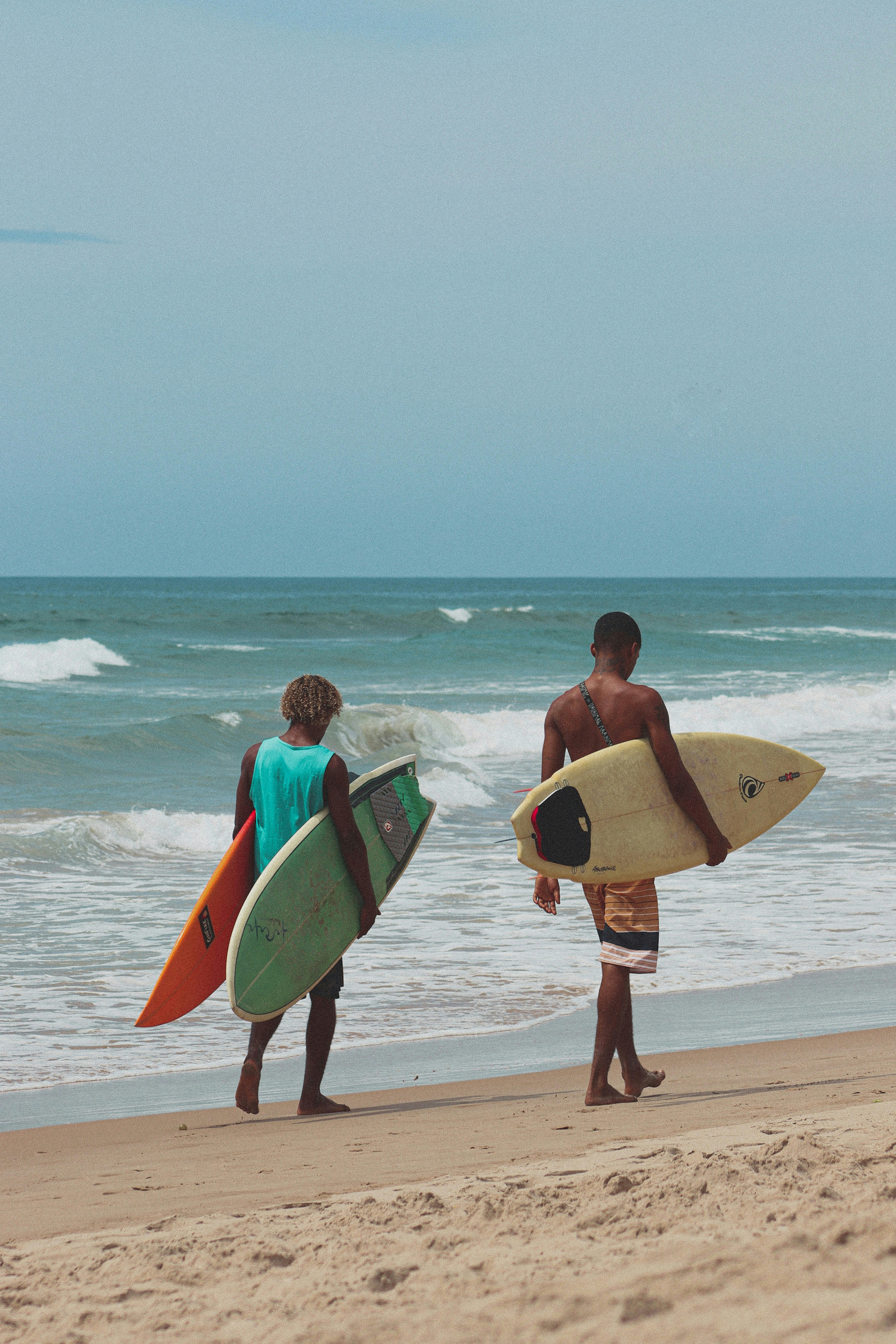 Two surfers walking along the beach, each carrying their surfboards as they head towards the ocean. The tranquil sea and soft sand create a relaxed atmosphere.