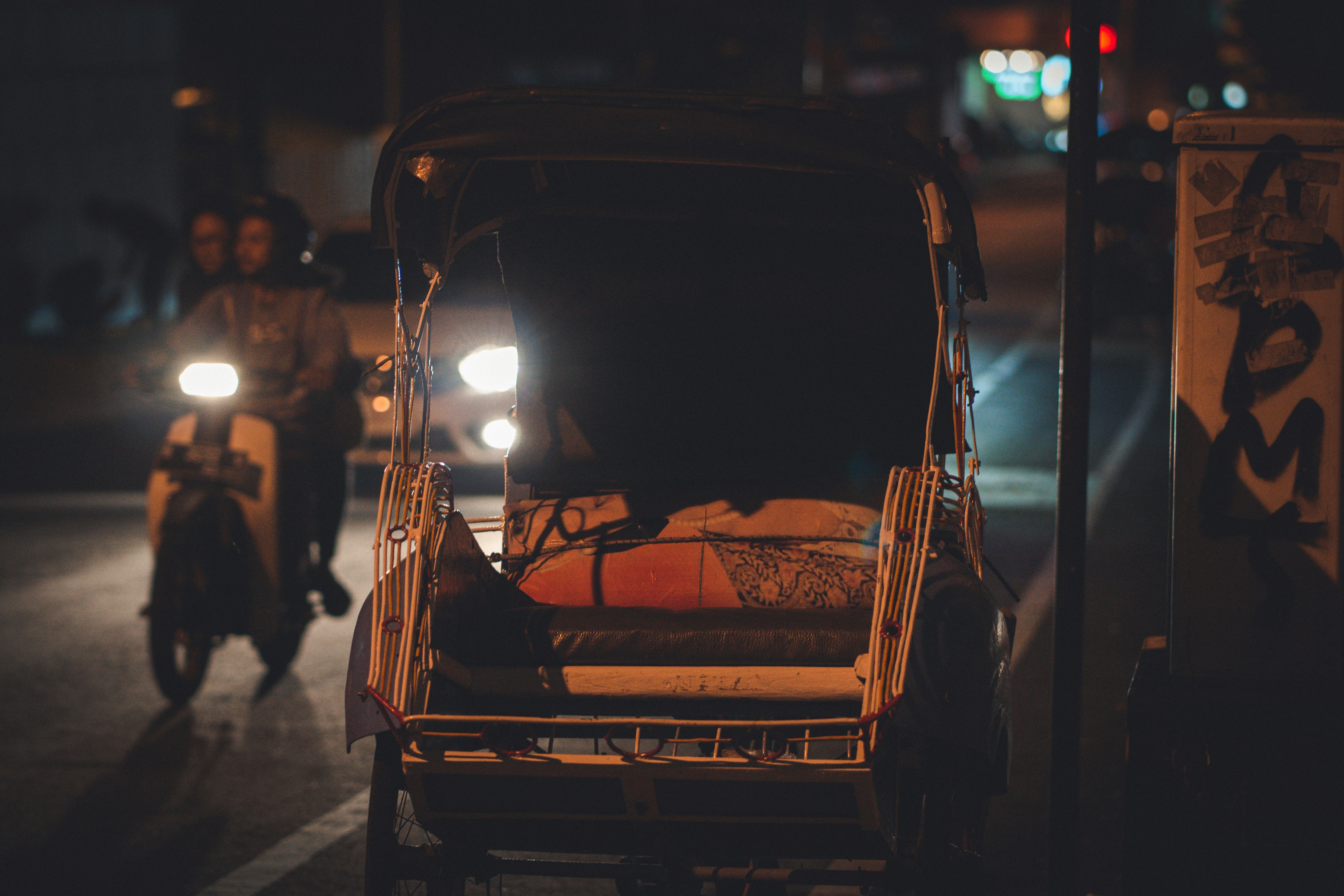 Rickshaw parked on a dimly lit street, with motorcyclists passing by in the background. The scene captures the essence of nighttime urban activity.