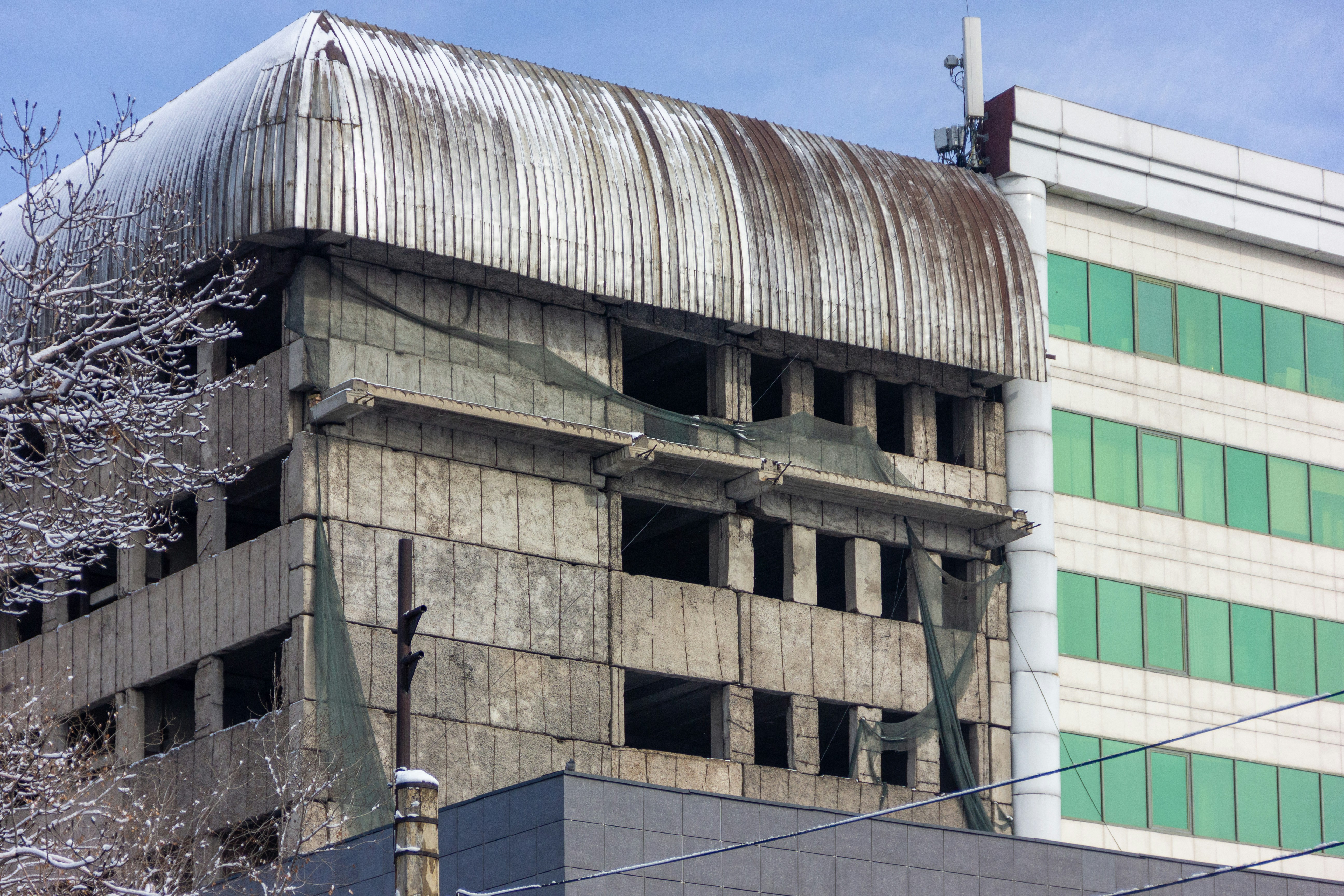 Dilapidated building with a rusted roof and exposed concrete, juxtaposed against a modern structure, highlighting urban decay.