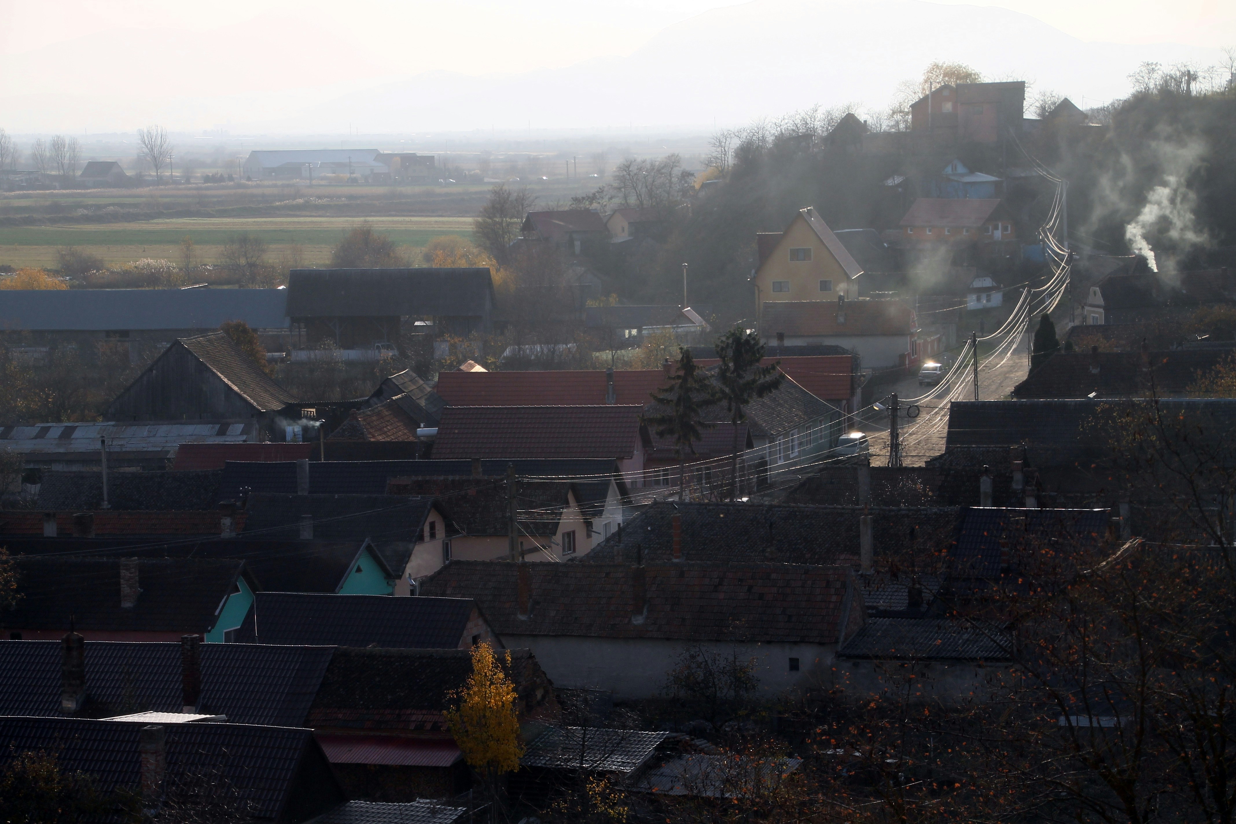 A serene view of a village nestled among rolling hills, with smoke gently rising from chimneys and colorful rooftops peeking through the mist.