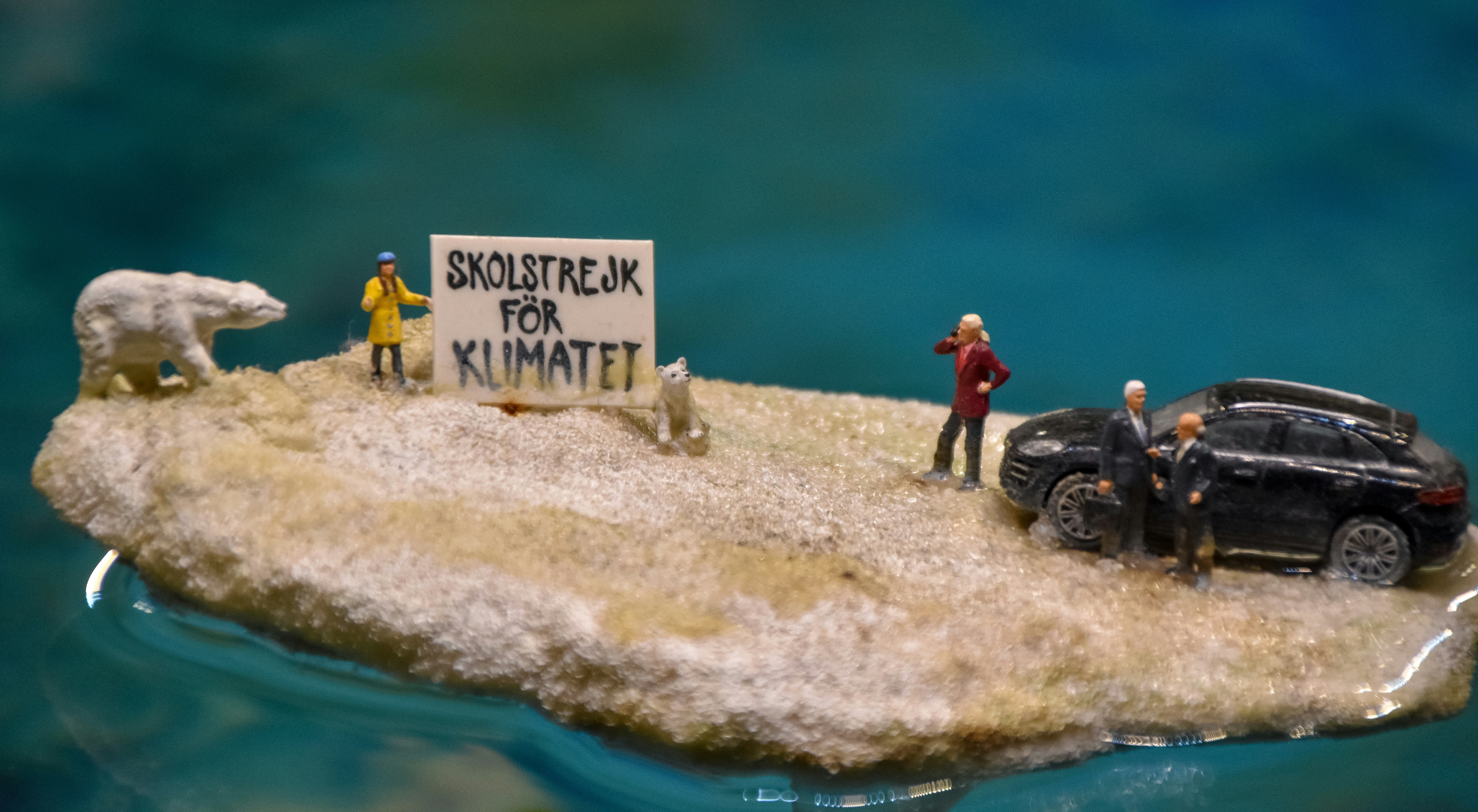 Miniature figures on a small island hold a sign advocating for climate action, surrounded by a polar bear and a sleek black vehicle.