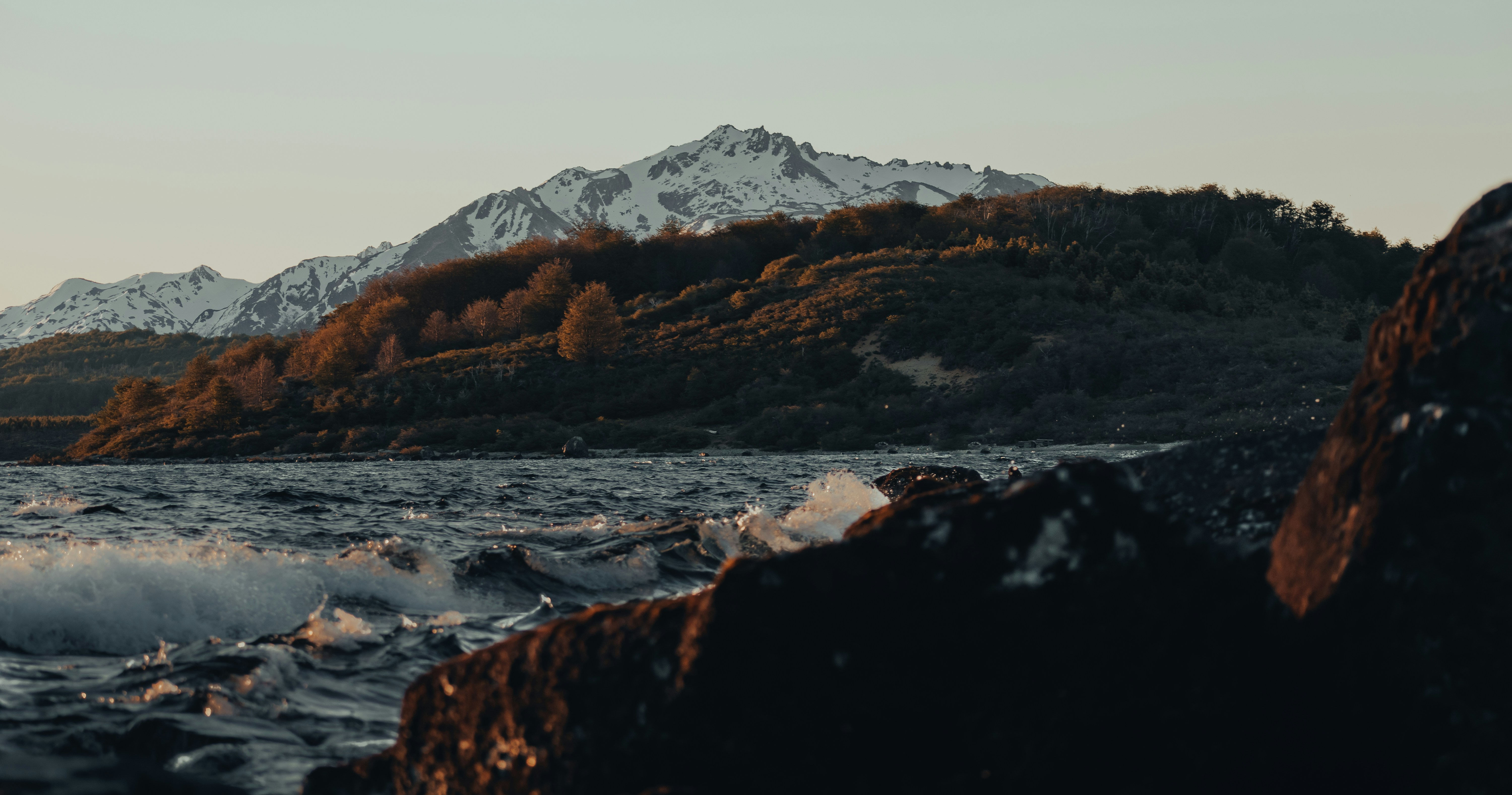 Waves lapping against dark rocks with a backdrop of snow-capped mountains and lush hills under a soft evening light.
