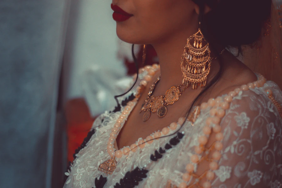 Close-up of a woman in bridal attire wearing ornate gold earrings, a necklace, and a white lace blouse with pearl trim. Her red lipstick and jeweled details stand out against the softly blurred background.