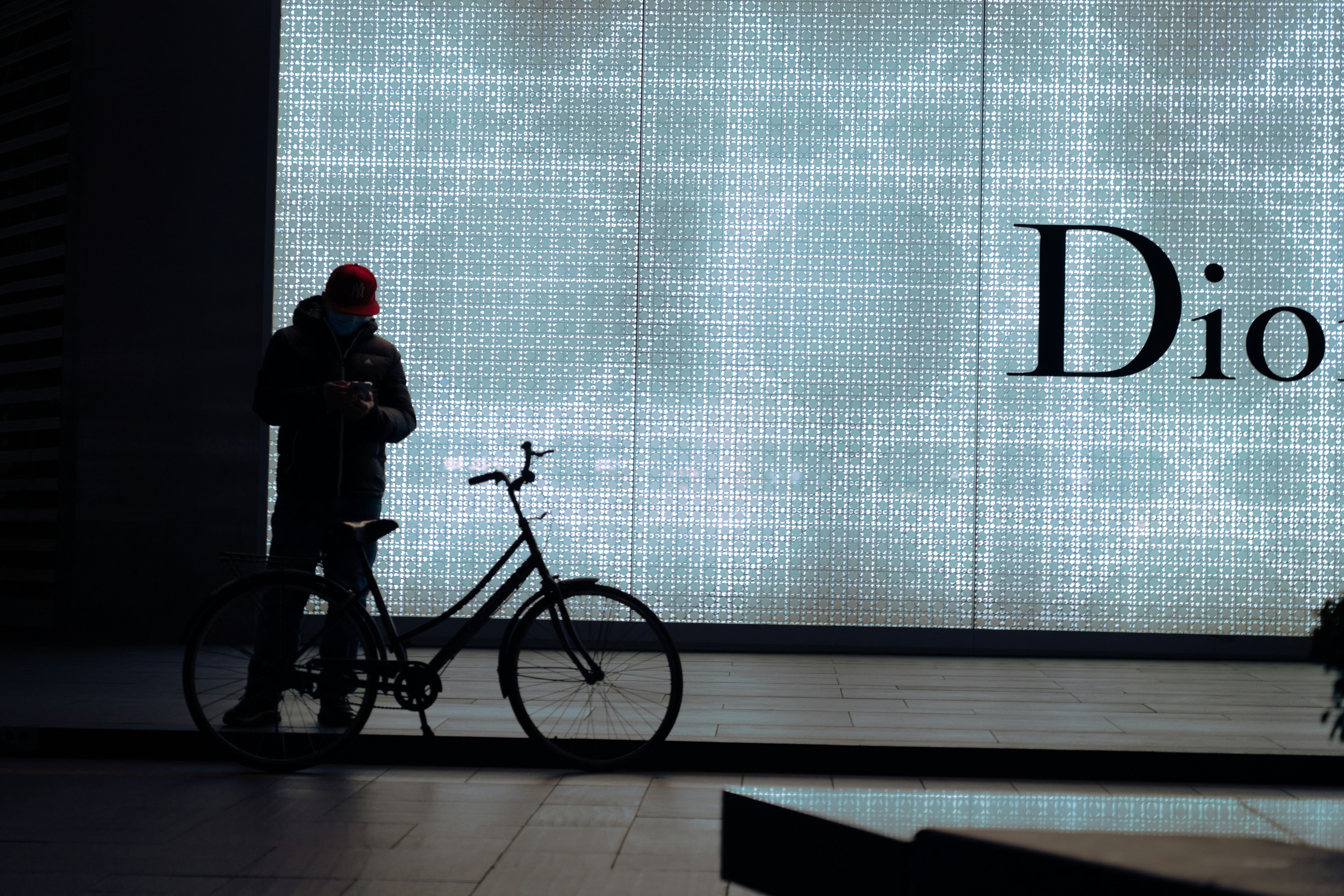 A lone figure with a red cap stands beside a bicycle, engrossed in a mobile device against a vibrant digital backdrop featuring the word 'Dior.'