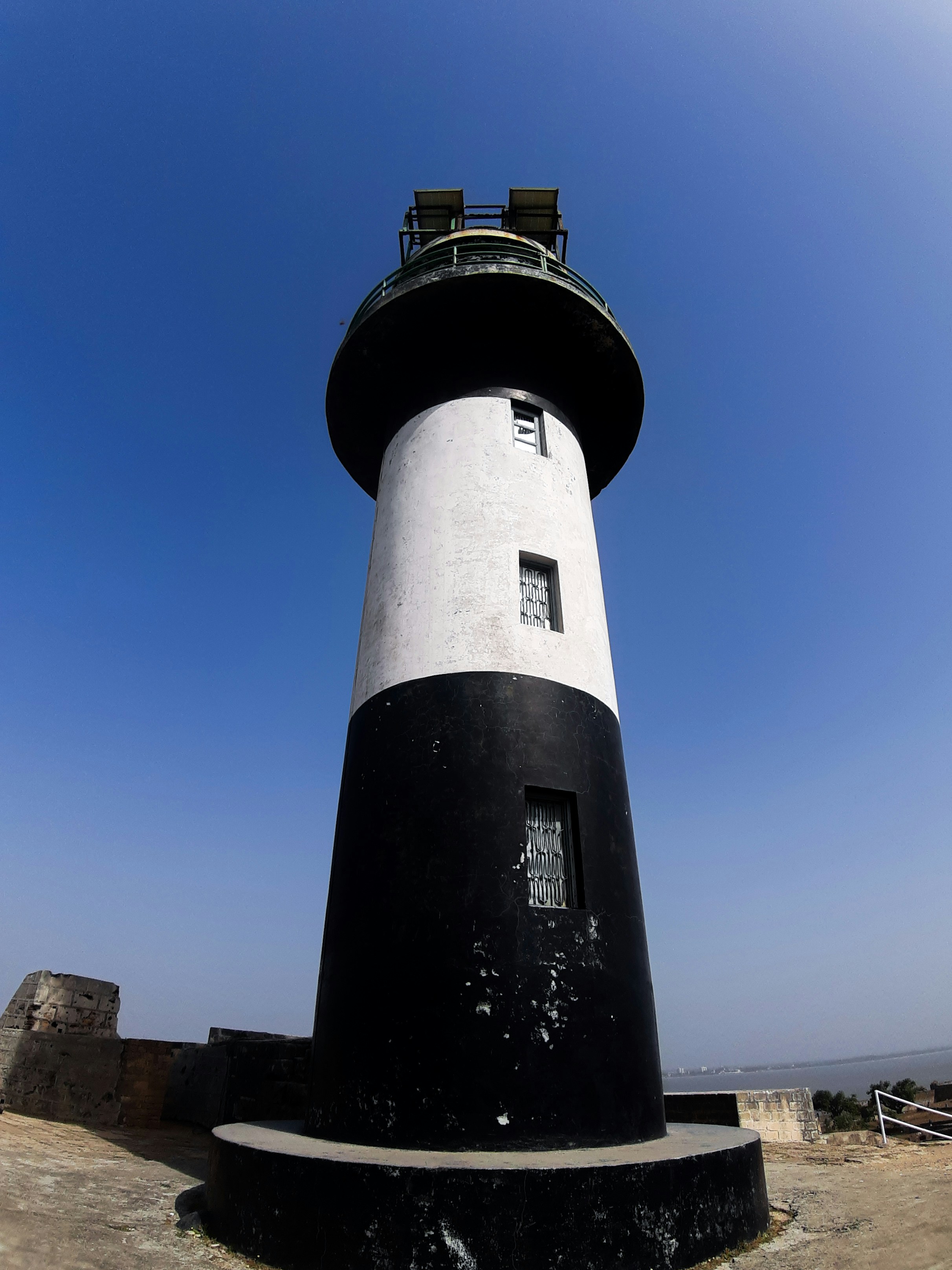 Two-tone lighthouse with a black lower band and white upper section rises against a clear blue sky. The stark tower anchors the coastal scene, with narrow windows and a gallery at the top.