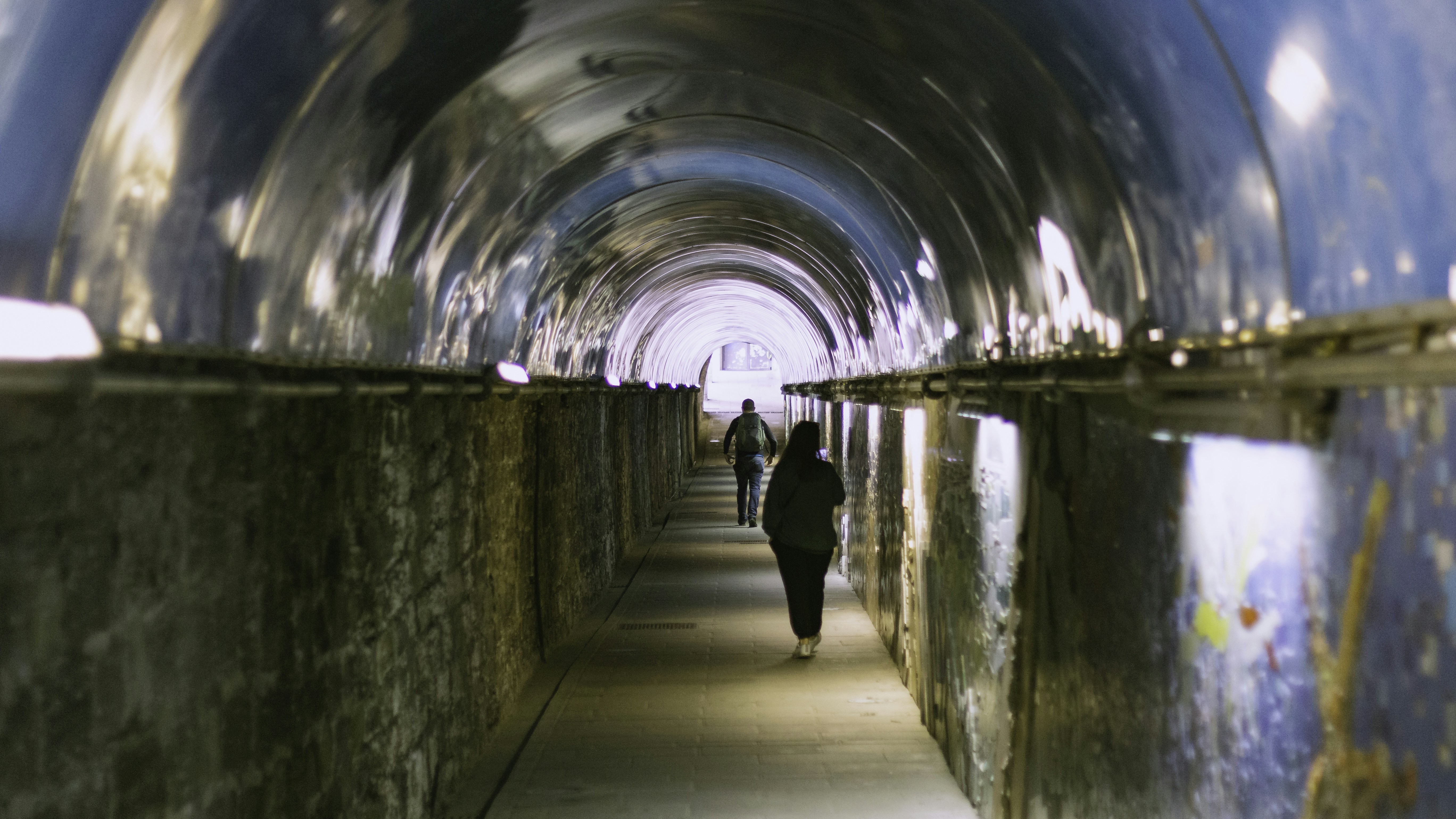 Two figures walking through a dimly lit tunnel, showcasing the contrast between light and shadow in an urban environment.