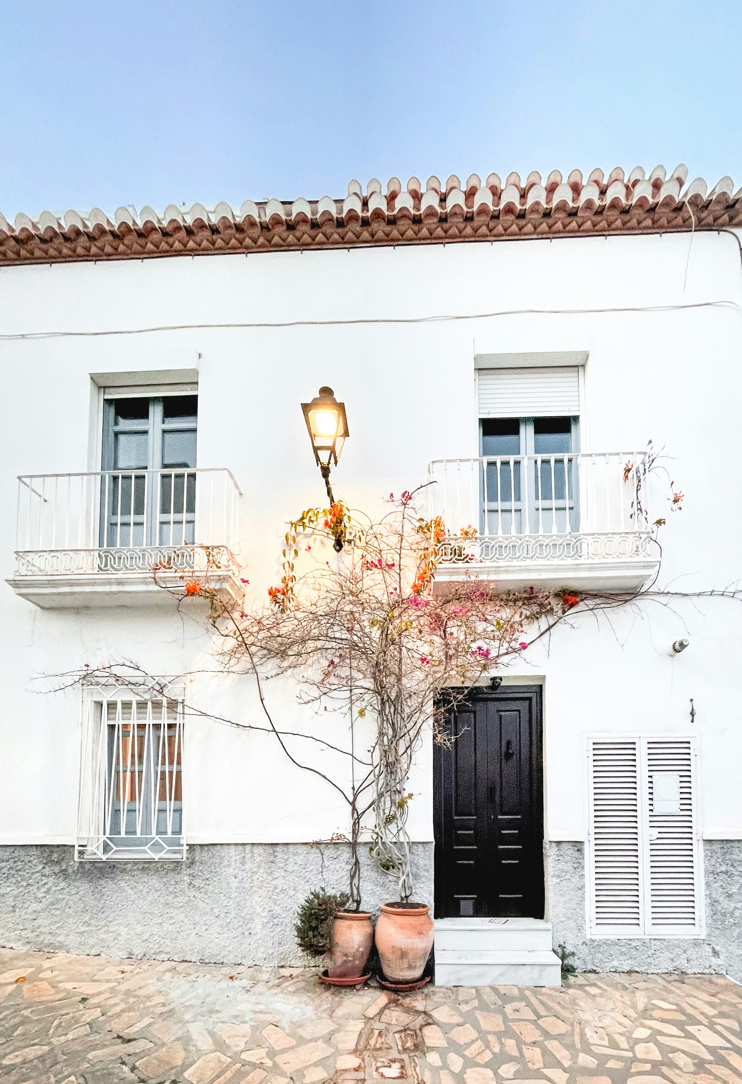 Whitewashed house with potted plants, wrought iron balconies, and central lamppost under a clear blue sky.
