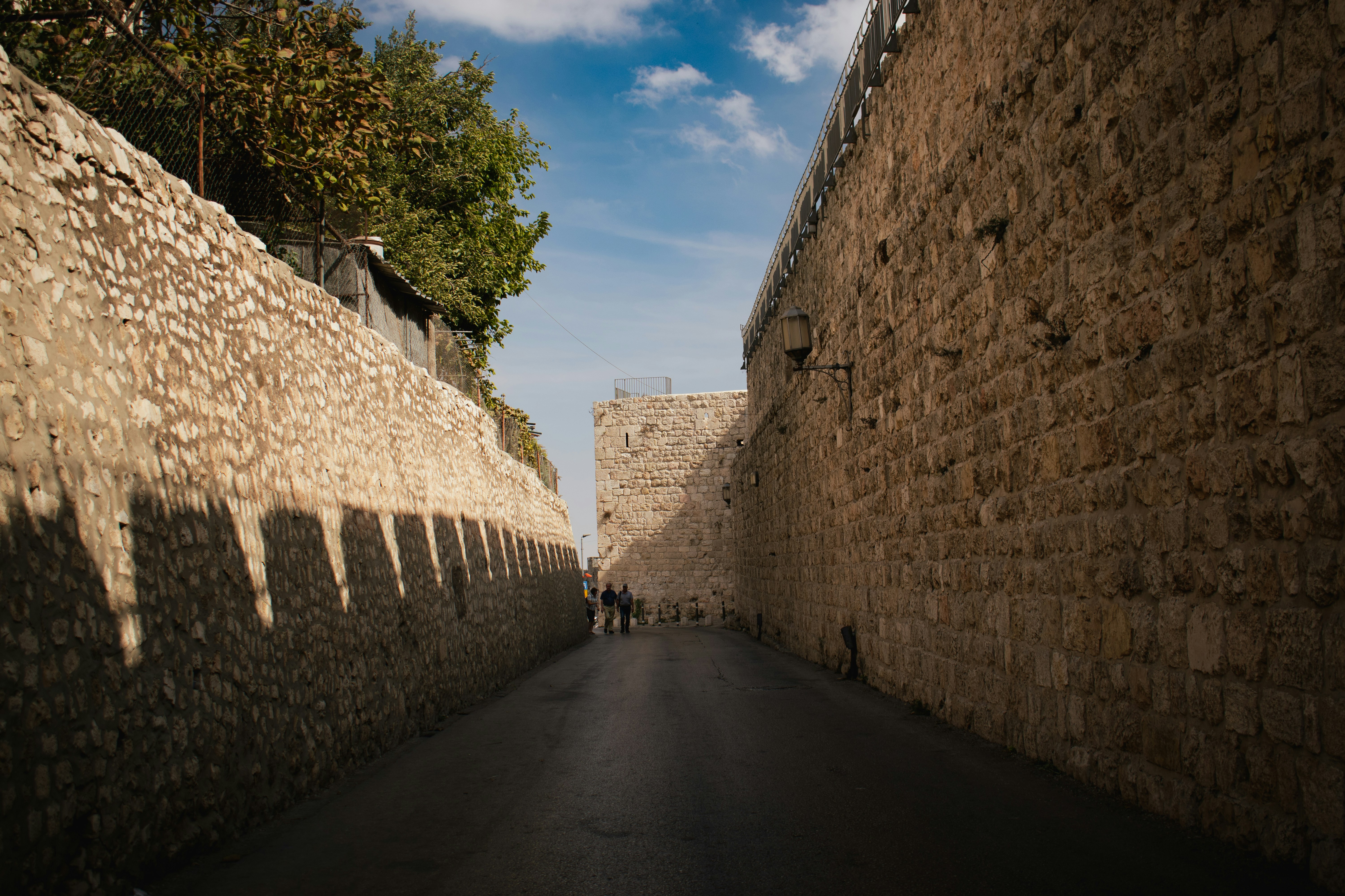 Narrow stone pathway flanked by ancient walls under a bright sky, with figures walking in the distance. The scene captures a moment of exploration in a historic setting.