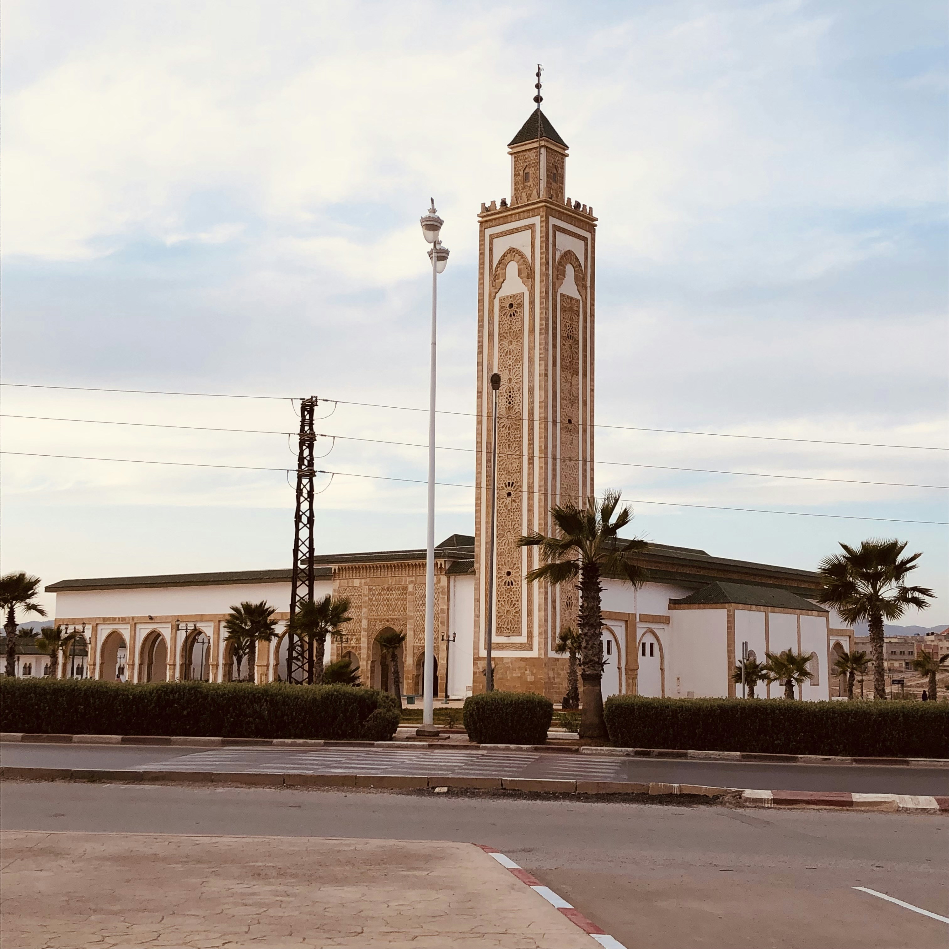 Tall minaret of a mosque against a serene sky, surrounded by palm trees and quiet streets.