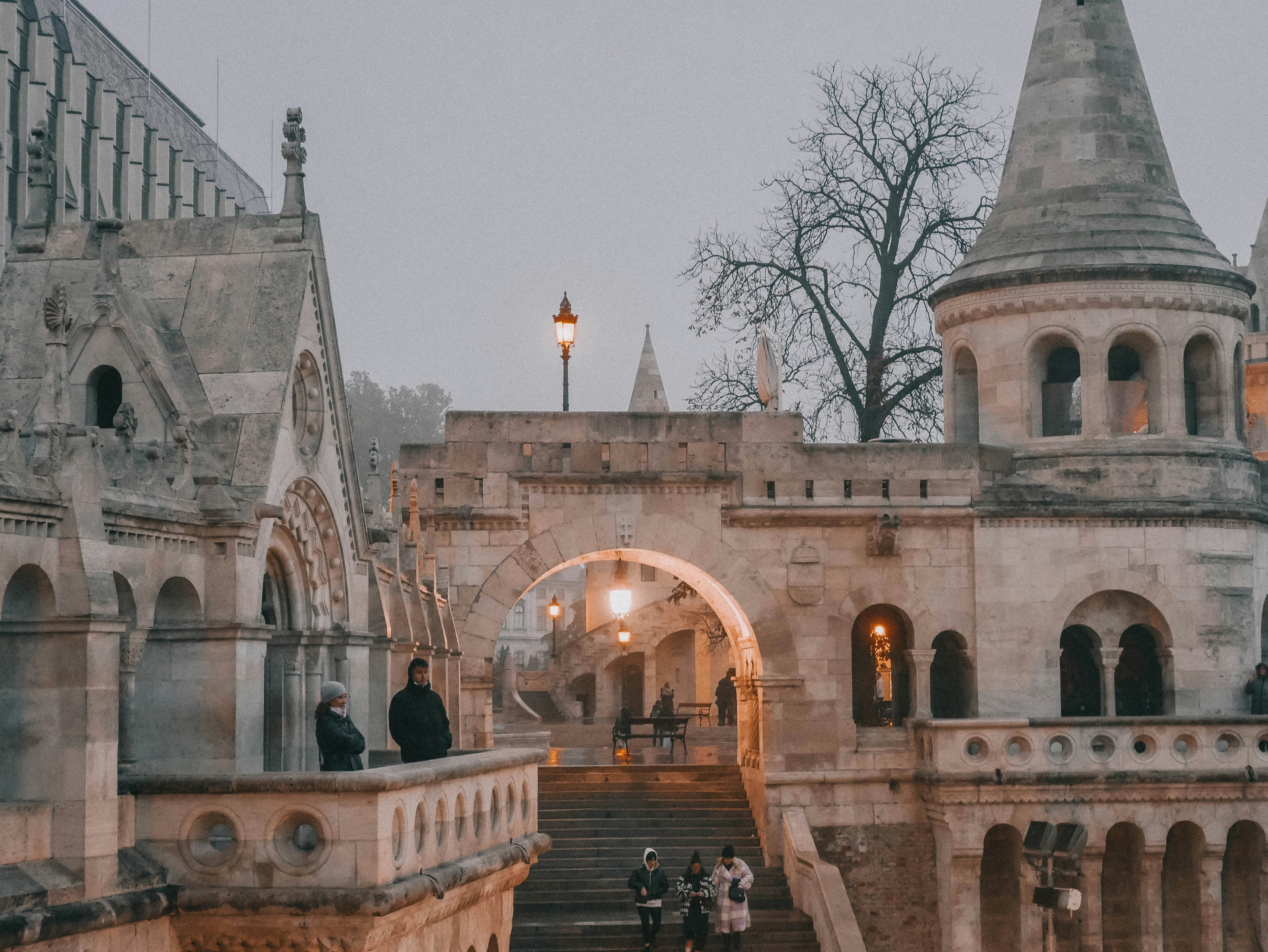 Stone archway fortress at dusk with warm lantern light along a stone stairway and a few silhouetted figures, highlighting architectural rhythm and texture.
