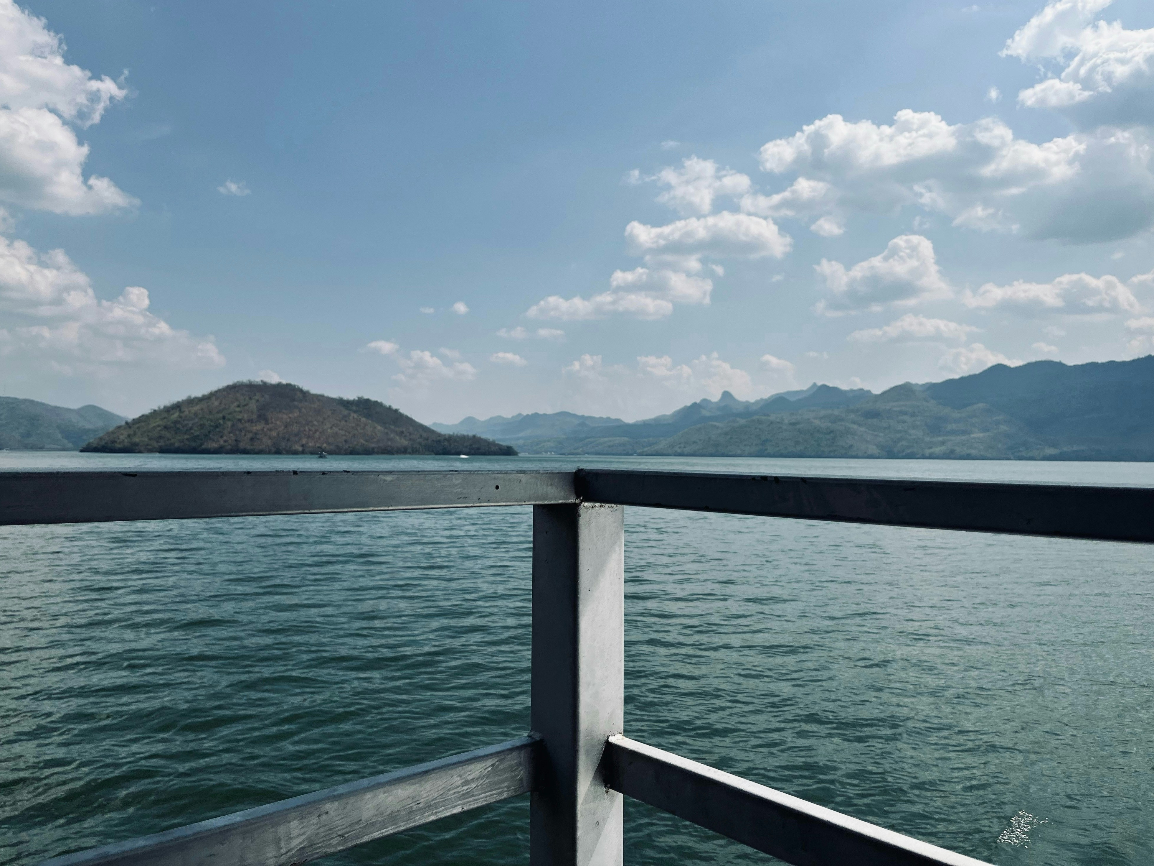 A tranquil view from a dock, showcasing a calm lake and distant mountains under a clear sky with fluffy clouds.