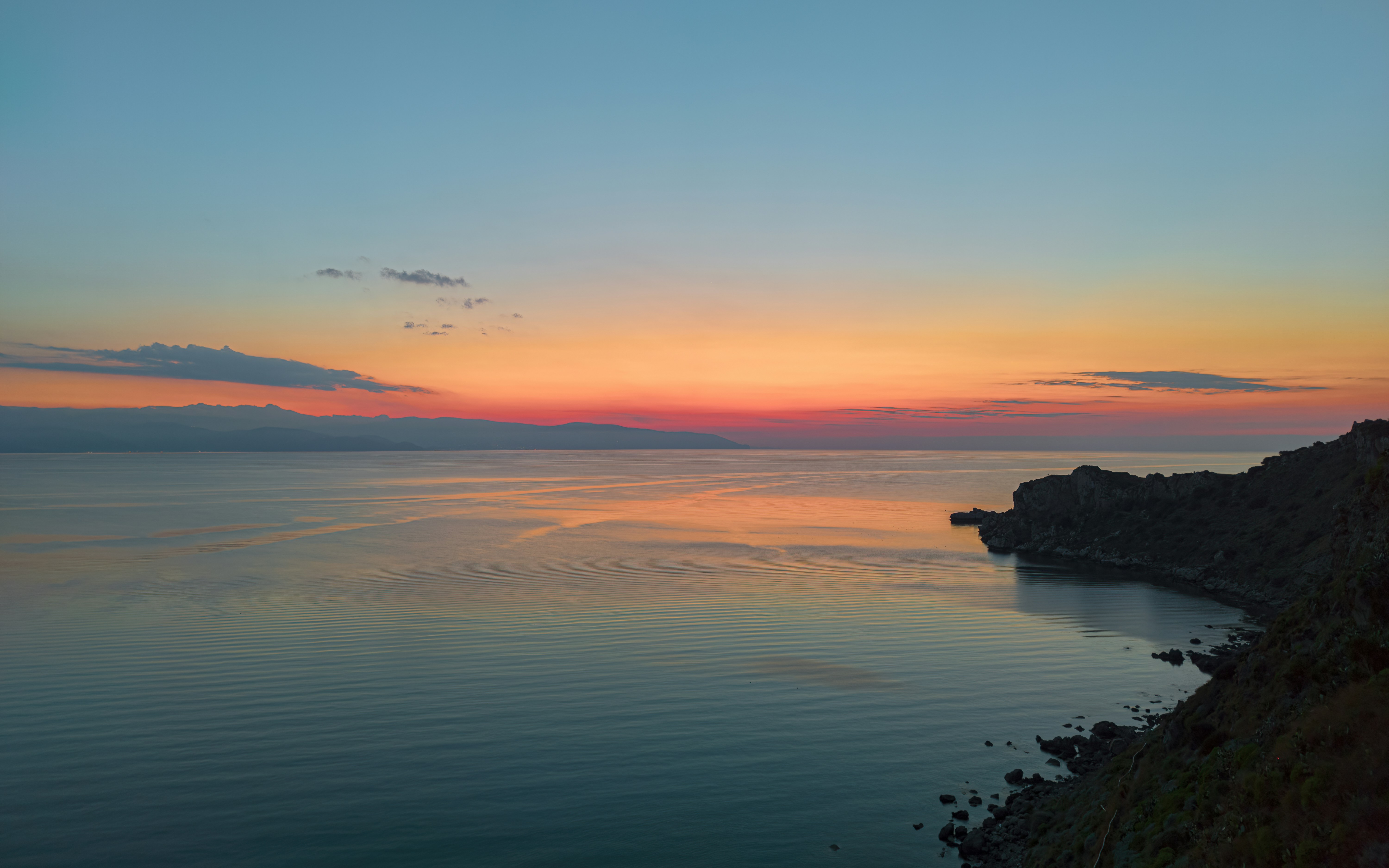 Vibrant sunset reflecting on calm waters, framed by rocky coastline and distant mountains. The scene captures a tranquil moment of nature's beauty.