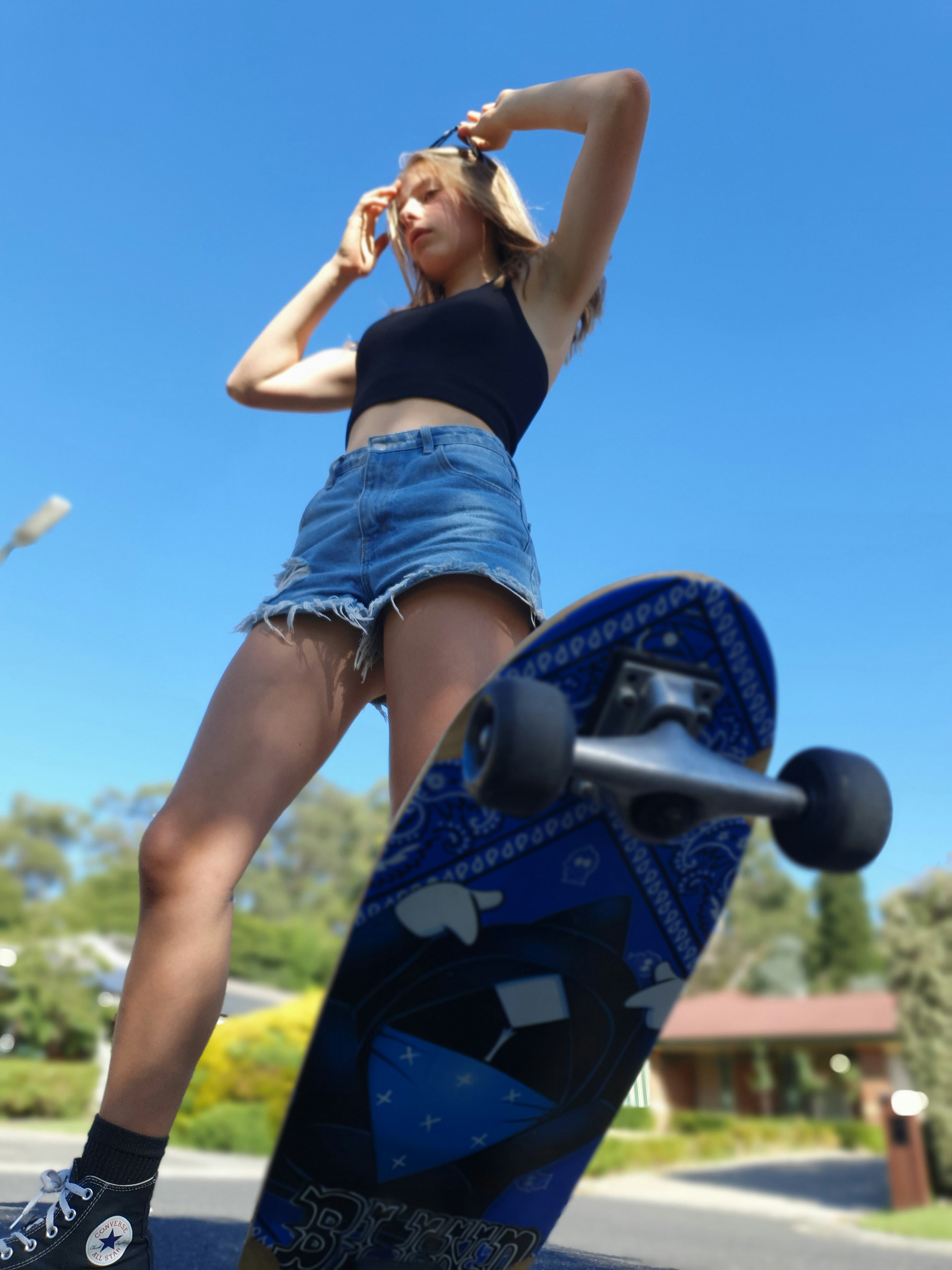 Young woman adjusting sunglasses while standing beside a skateboard on a sunny day. The vibrant blue sky enhances the scene's energy.