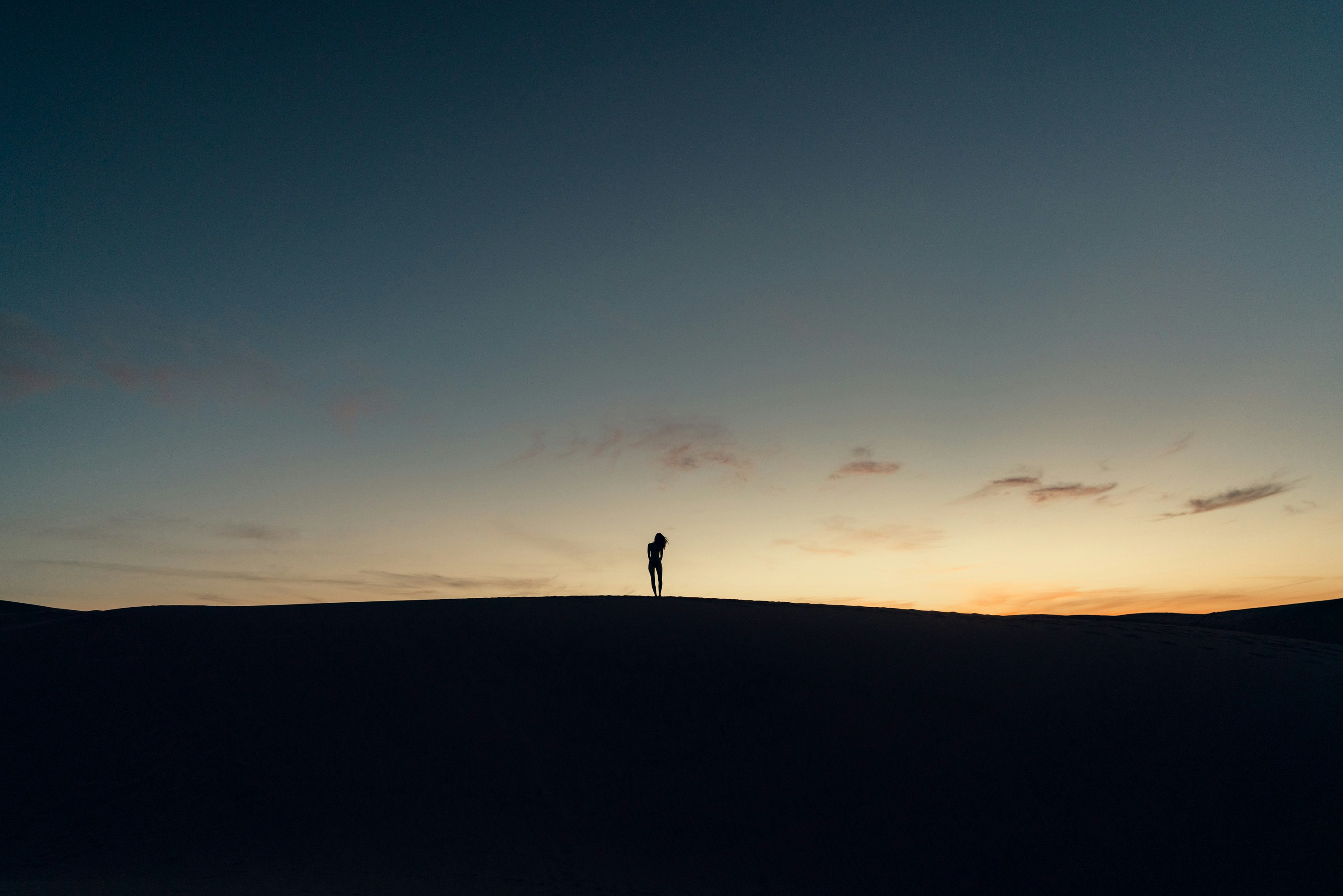 A lone silhouette stands atop a sand dune, framed by a gradient sky transitioning from dusk to night.