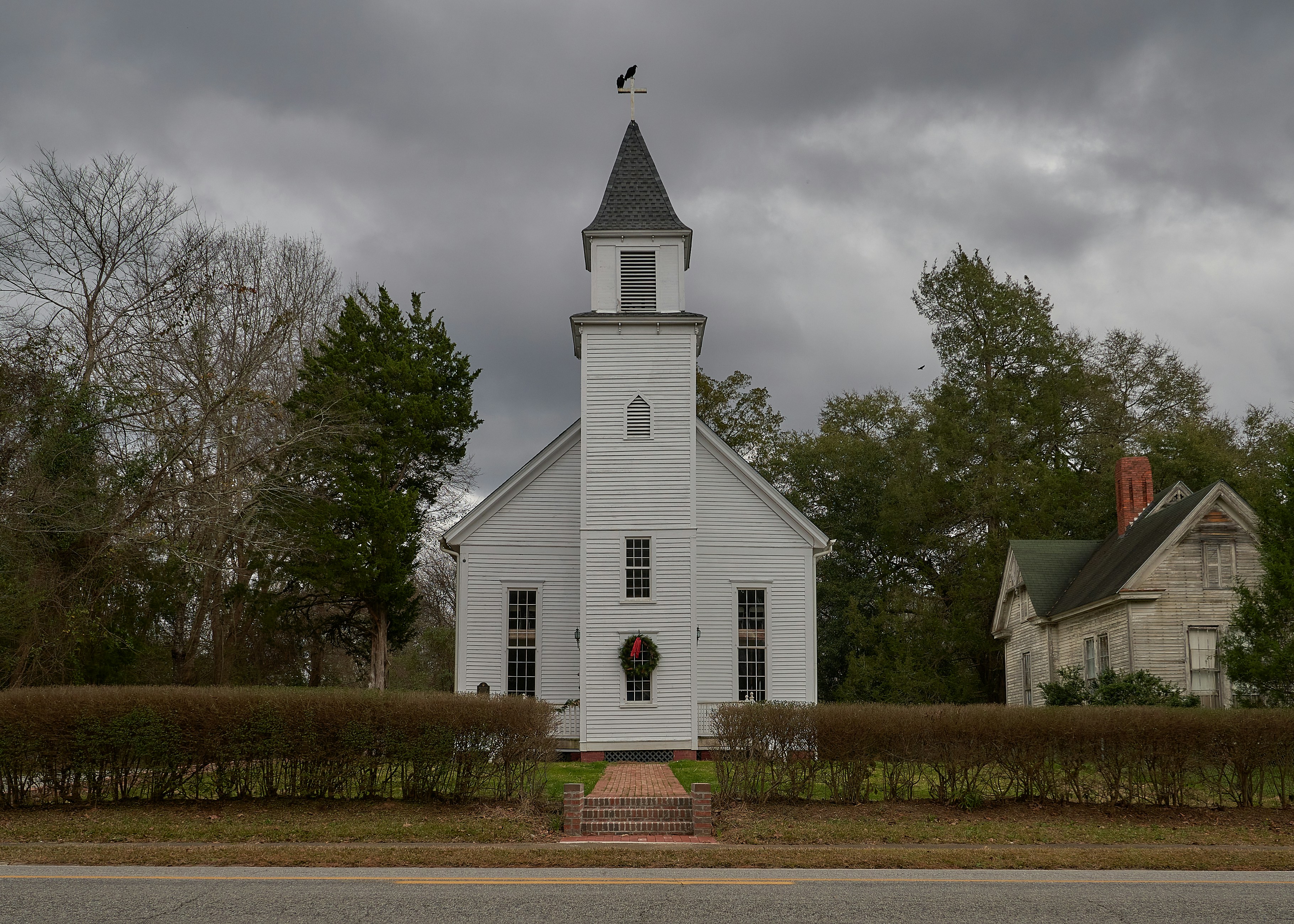 White chapel with a wreath on the door, flanked by trees under a cloudy sky.