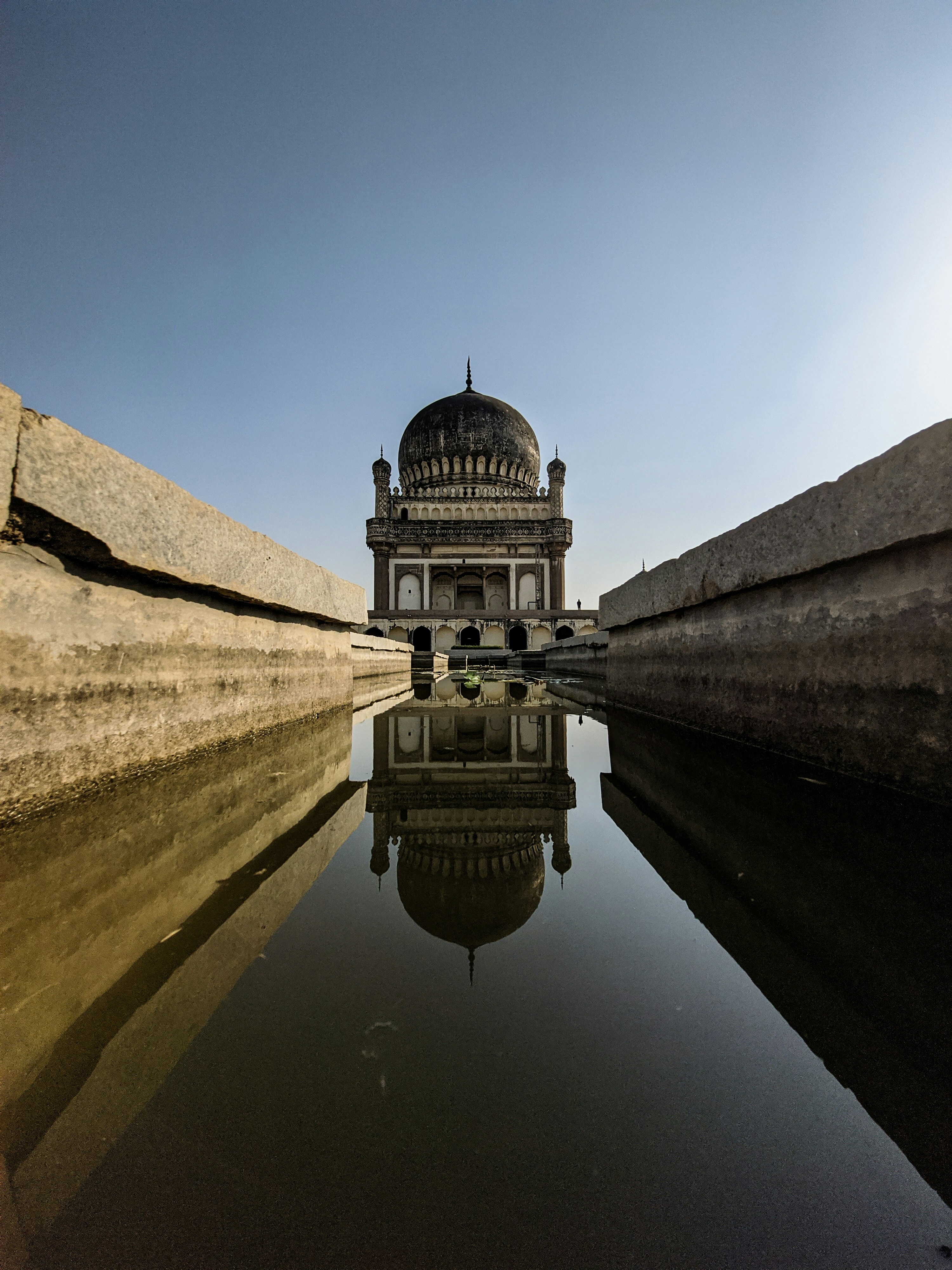Dome-topped mausoleum framed by stone banks with a calm water channel reflecting its image, creating a precise vertical symmetry.