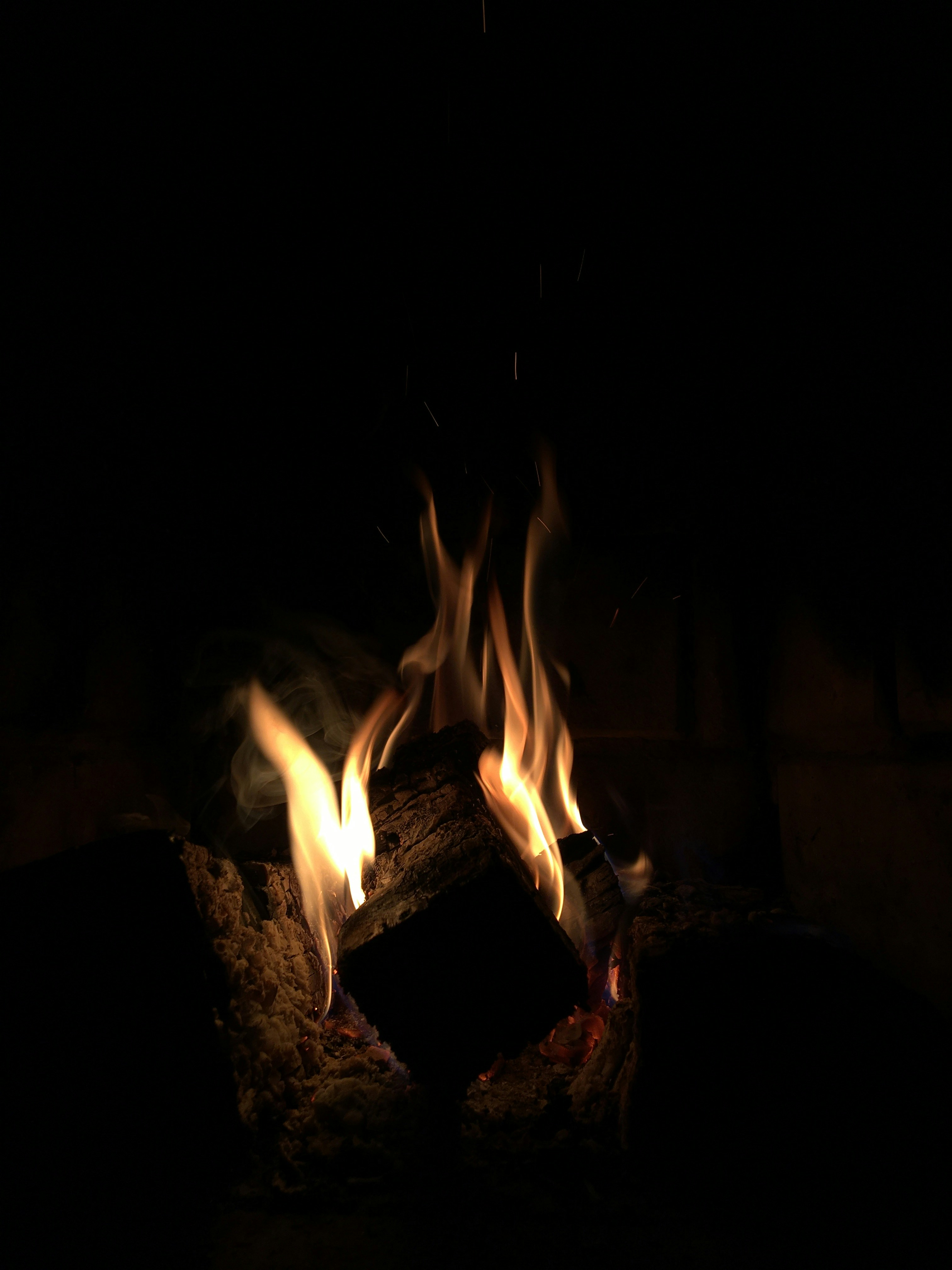 Flames flicker and swirl above glowing embers in a dark fireplace, creating a cozy atmosphere. The scene captures the essence of warmth and comfort.