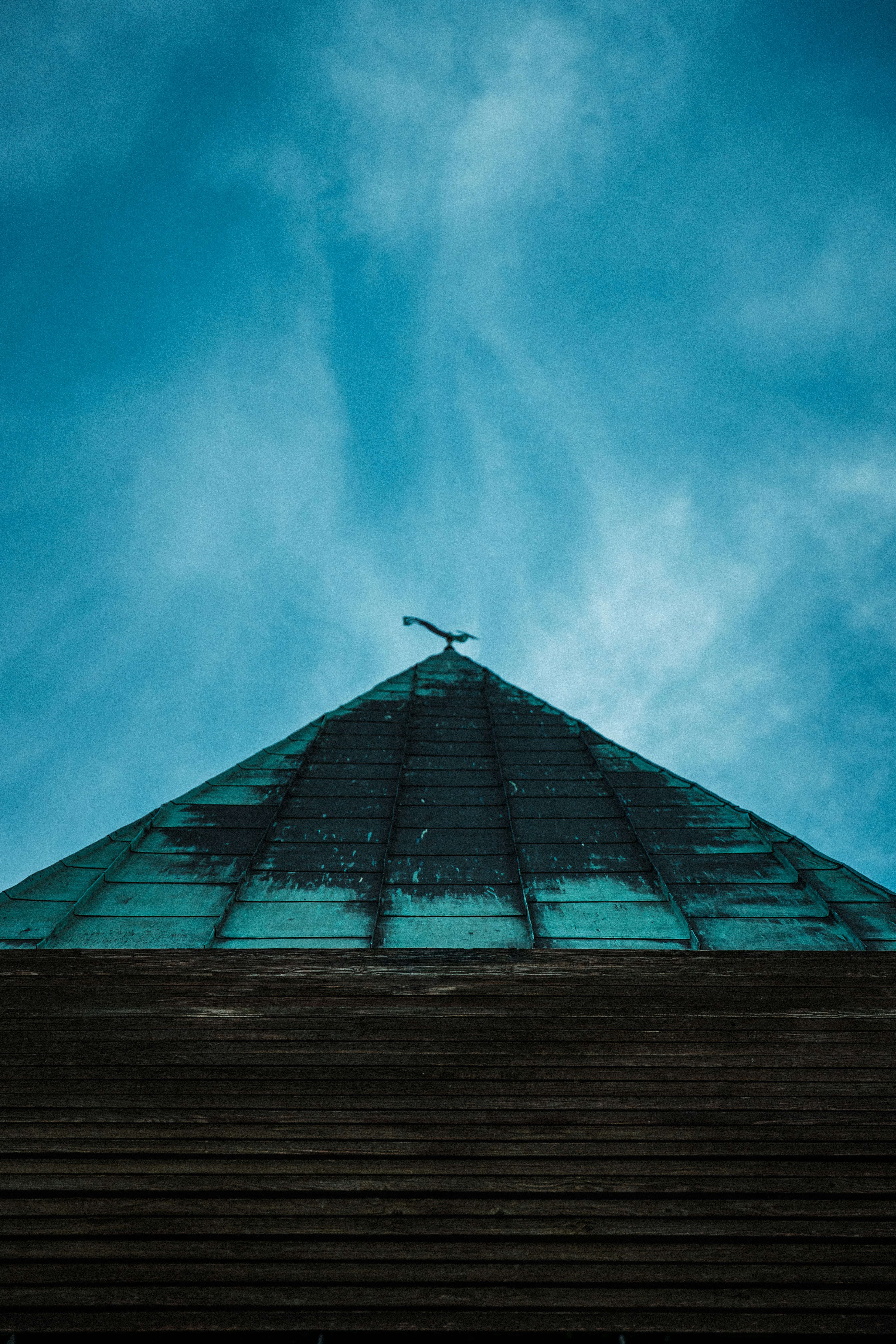 The pointed roof of a church tower rises against a dramatic blue sky, showcasing intricate textures and lines. A cross adorns the apex, symbolizing faith.