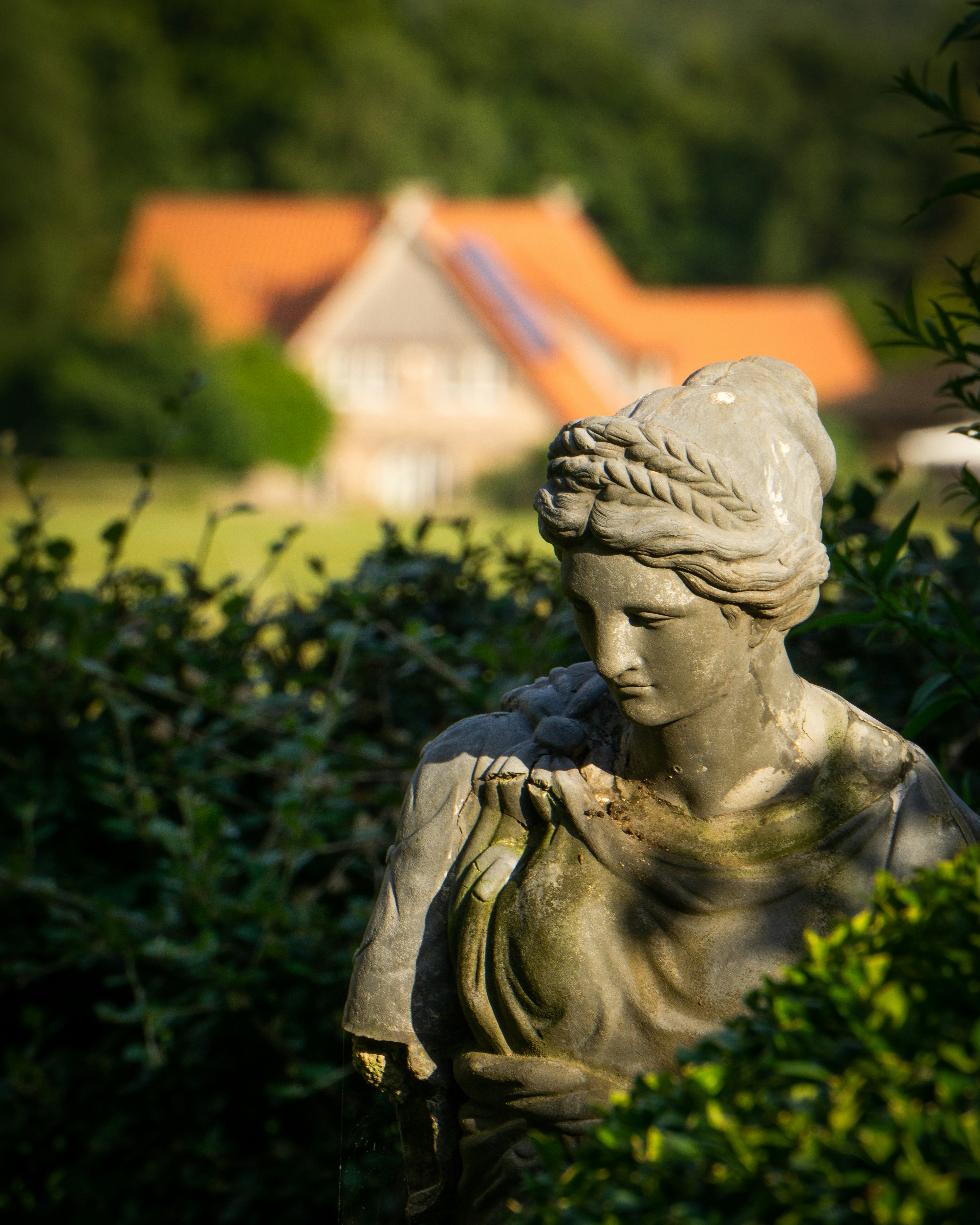 Stone Maiden in a Sunlit Garden, Framed by an Orange-Roofed House