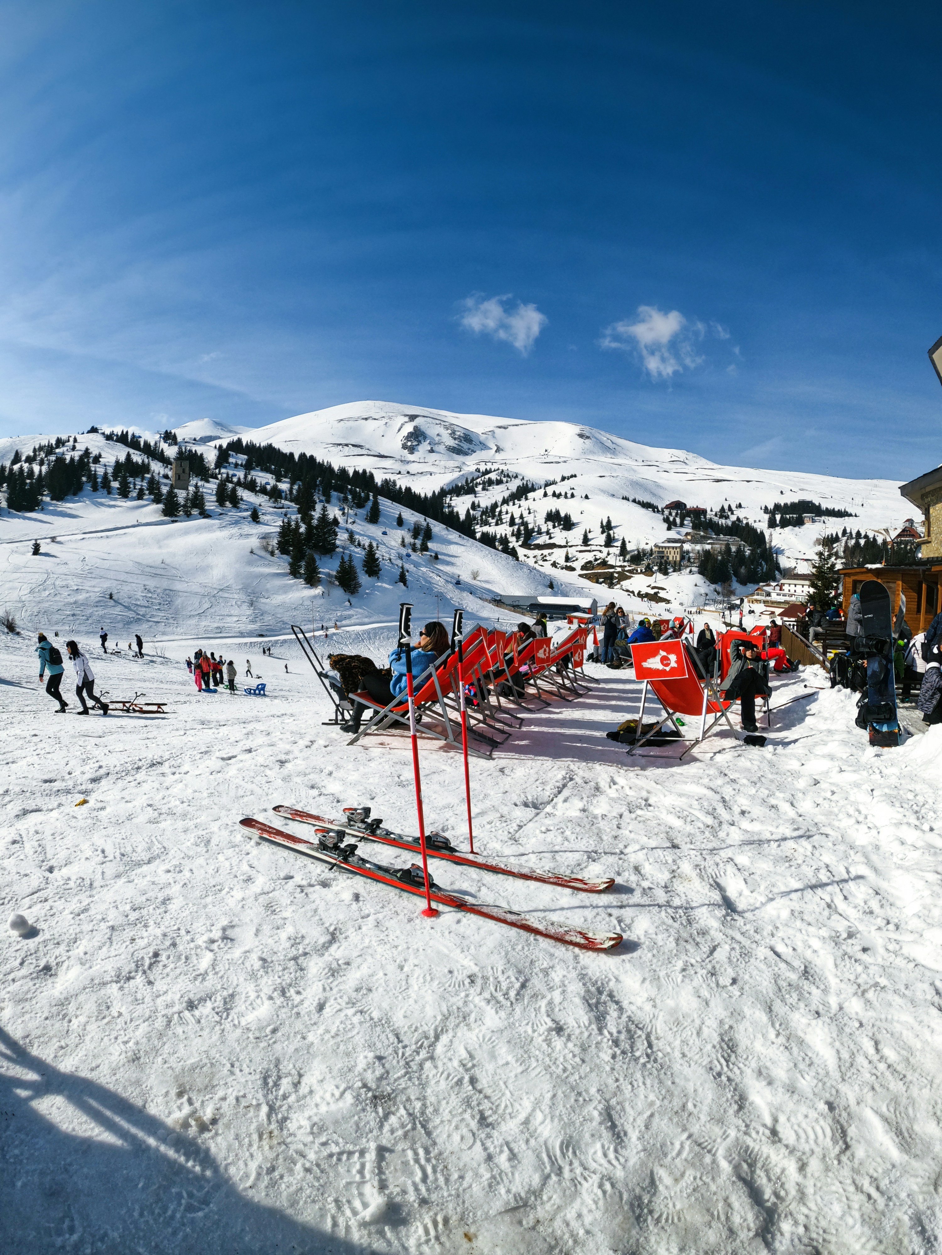 Snow-covered alpine village scene with a row of red ski chairs and skiers resting on a bright slope, framed by distant snow-clad mountains under a clear blue sky. A lively foreground of skis and visitors contrasts with the tranquil high peaks.