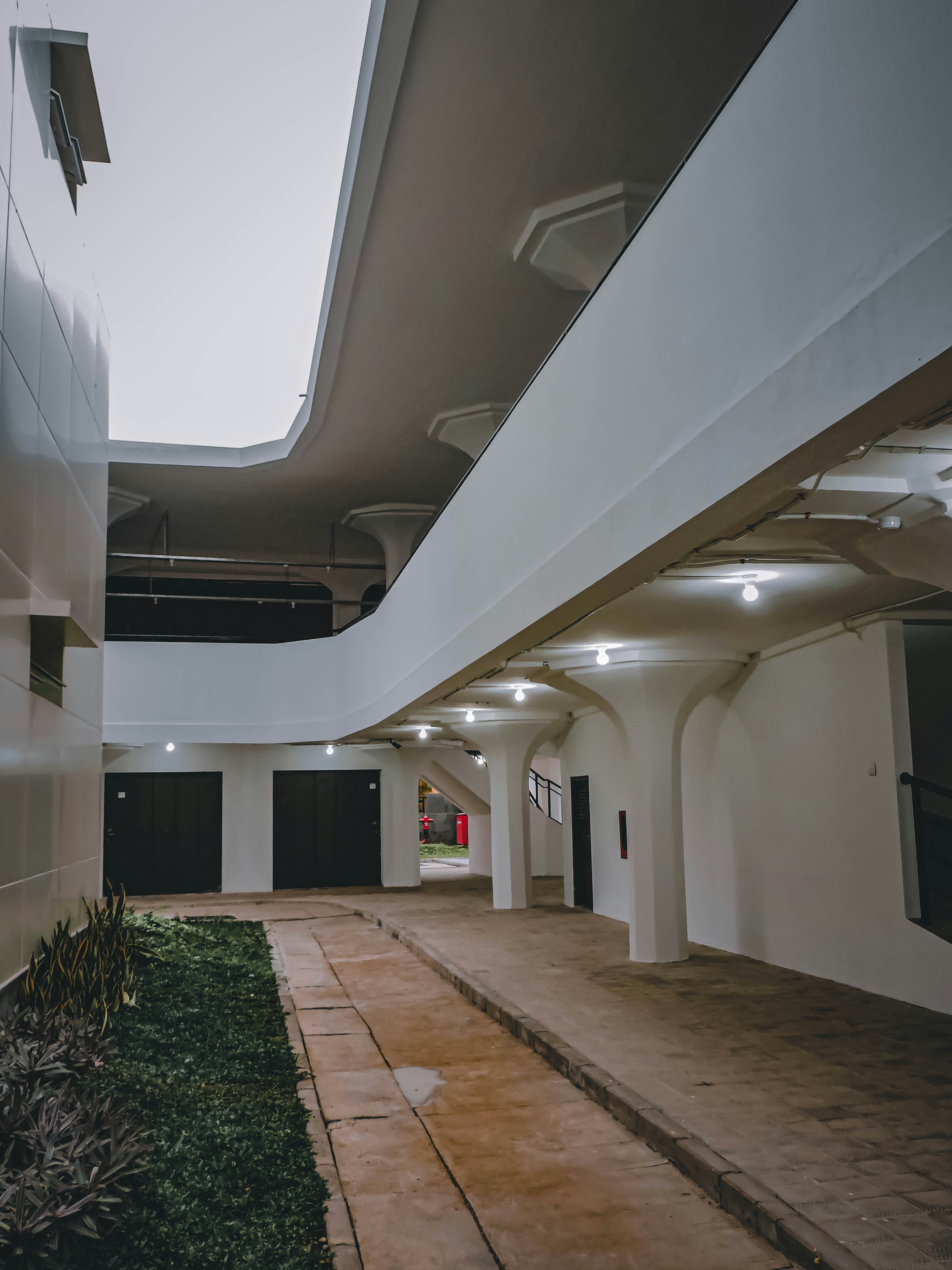 Architectural interior photograph featuring a white curved atrium, central walkway, and greenery along the left edge. Clean lines, arches, and soft overhead lighting define the space.