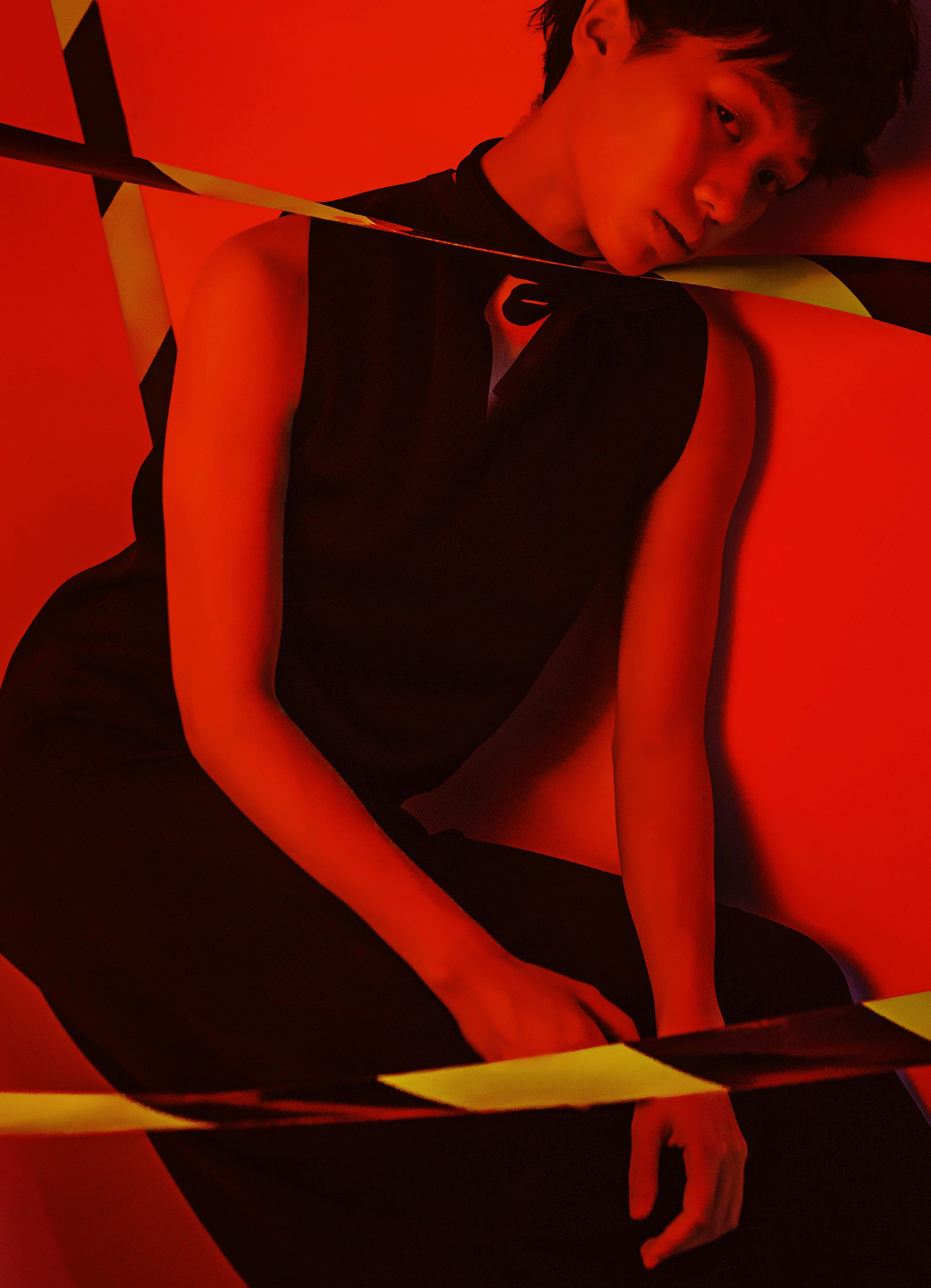 A figure in a black outfit leans against a wall, surrounded by yellow caution tape, set against a vibrant red background.