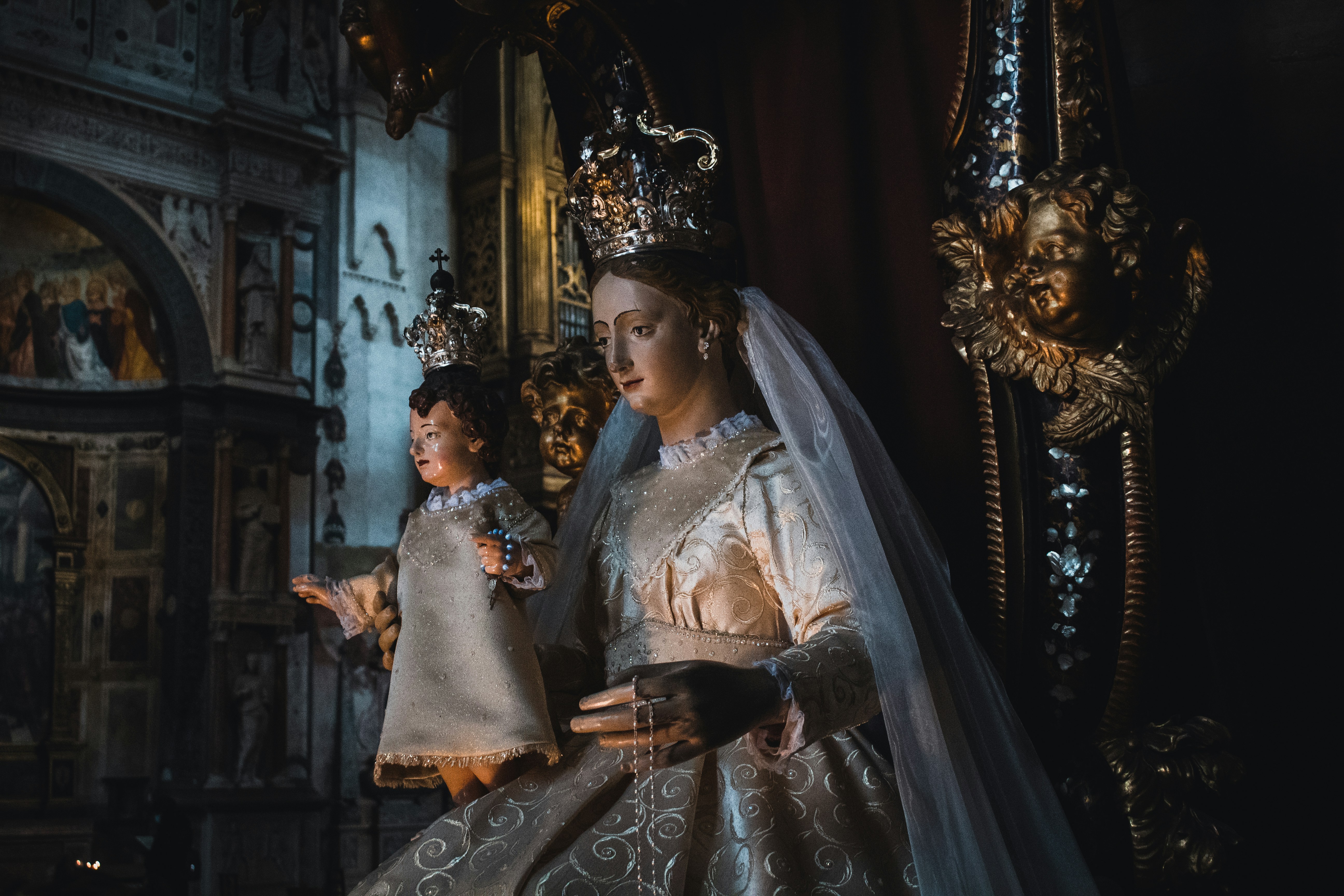 A beautifully adorned statue of a queenly figure holds a small doll, both crowned, illuminated softly against a richly detailed backdrop.