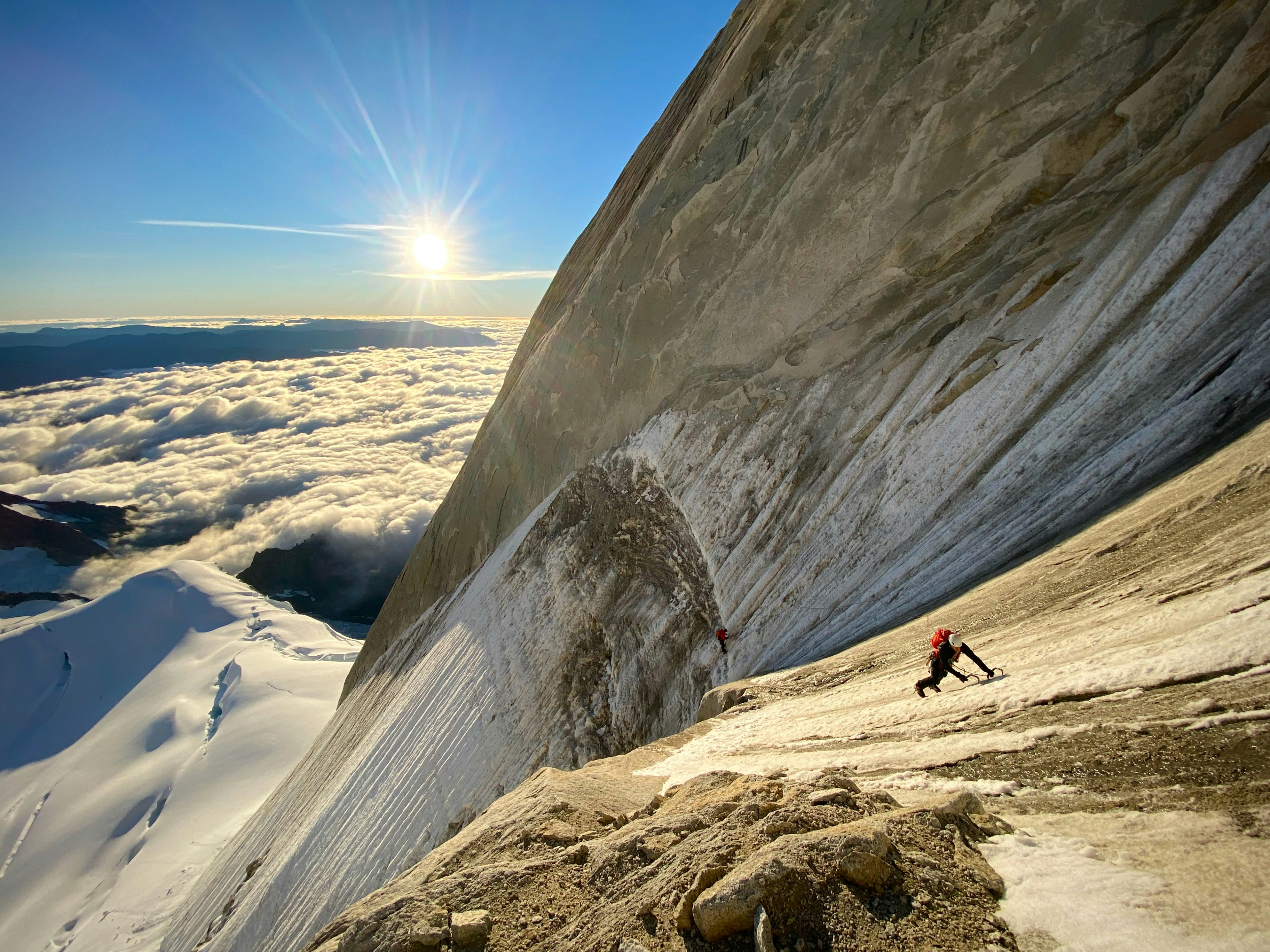 Climber navigating a steep, rocky face with a backdrop of clouds and a radiant sun setting on the horizon.